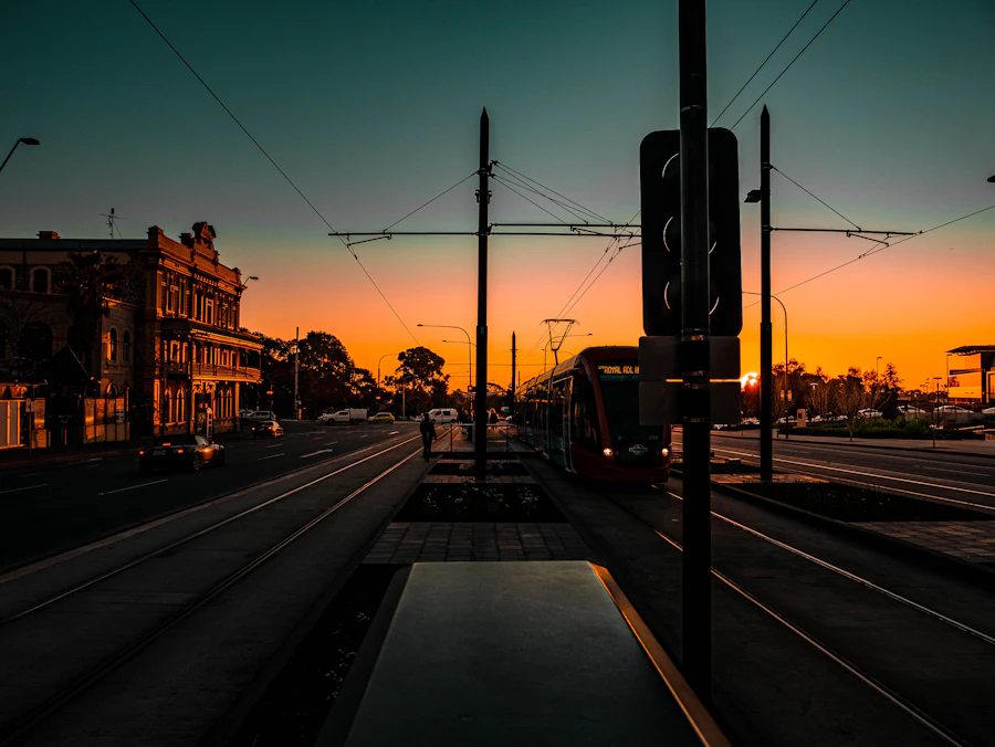 A tram waits along tracks at dusk, silhouetted against a vivid orange horizon. Dark buildings, poles, and overhead wires frame the scene.