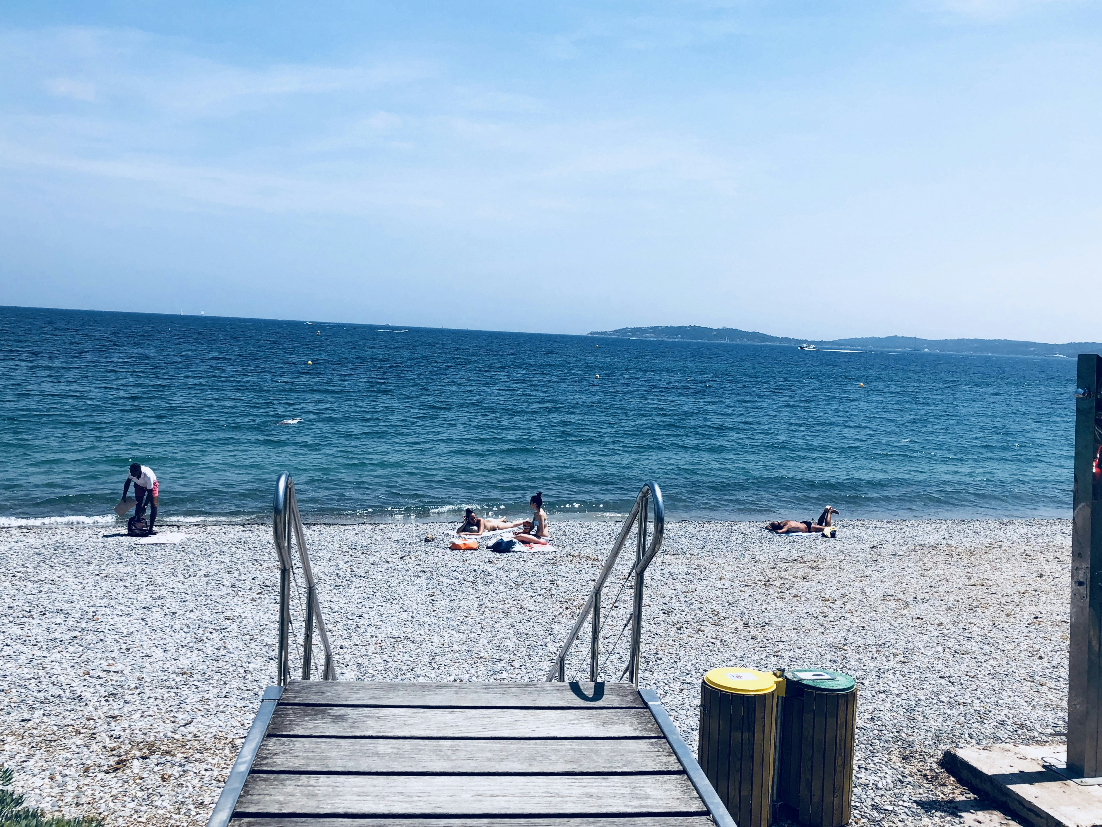Wooden boardwalk leading to a pebble beach with people relaxing under a clear blue sky.