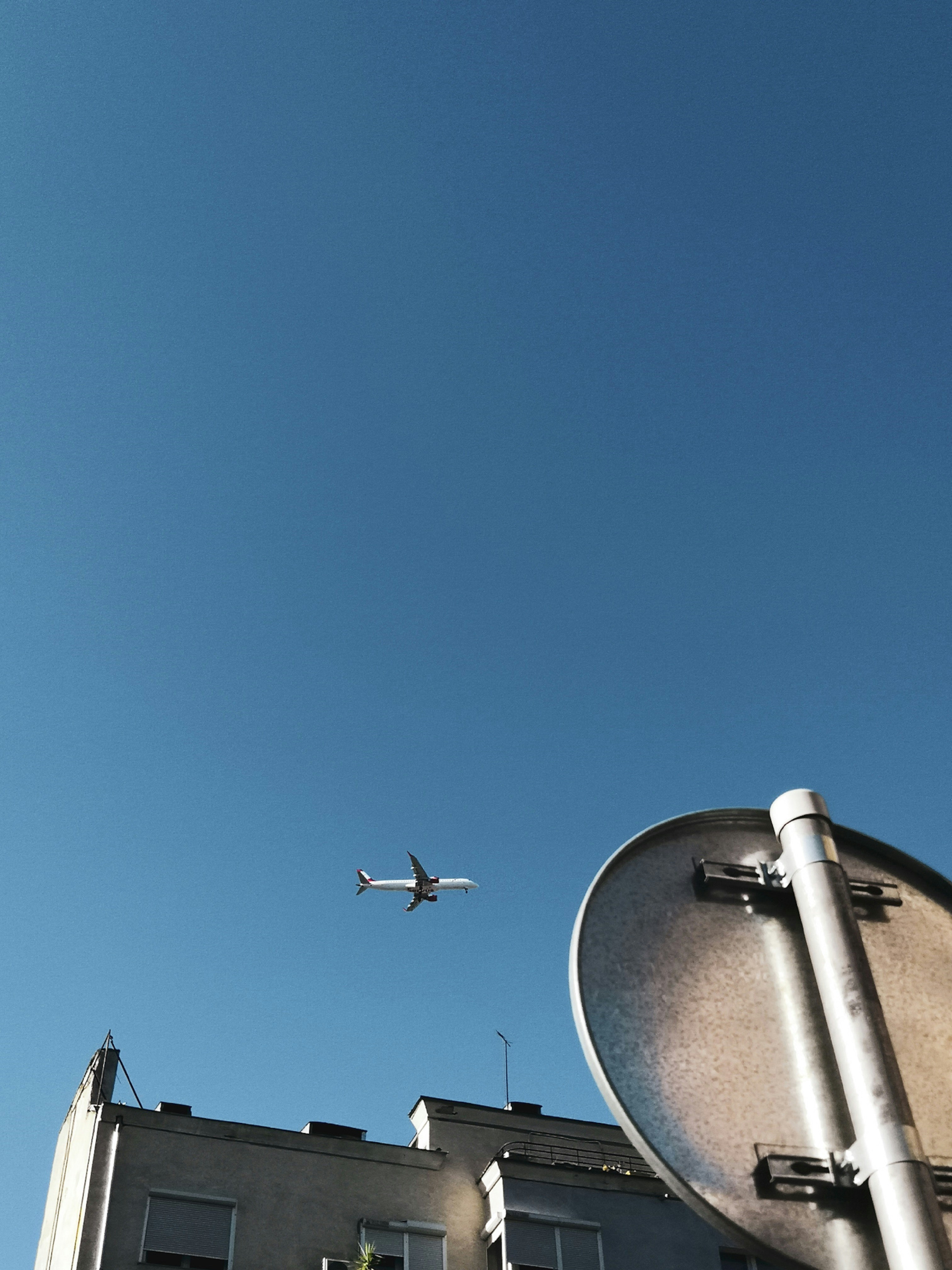 Photograph of a lone jet gliding across a clear blue sky above low city rooftops, with a round street sign occupying the foreground. The scene emphasizes scale and urban geometry against a vast, empty expanse.