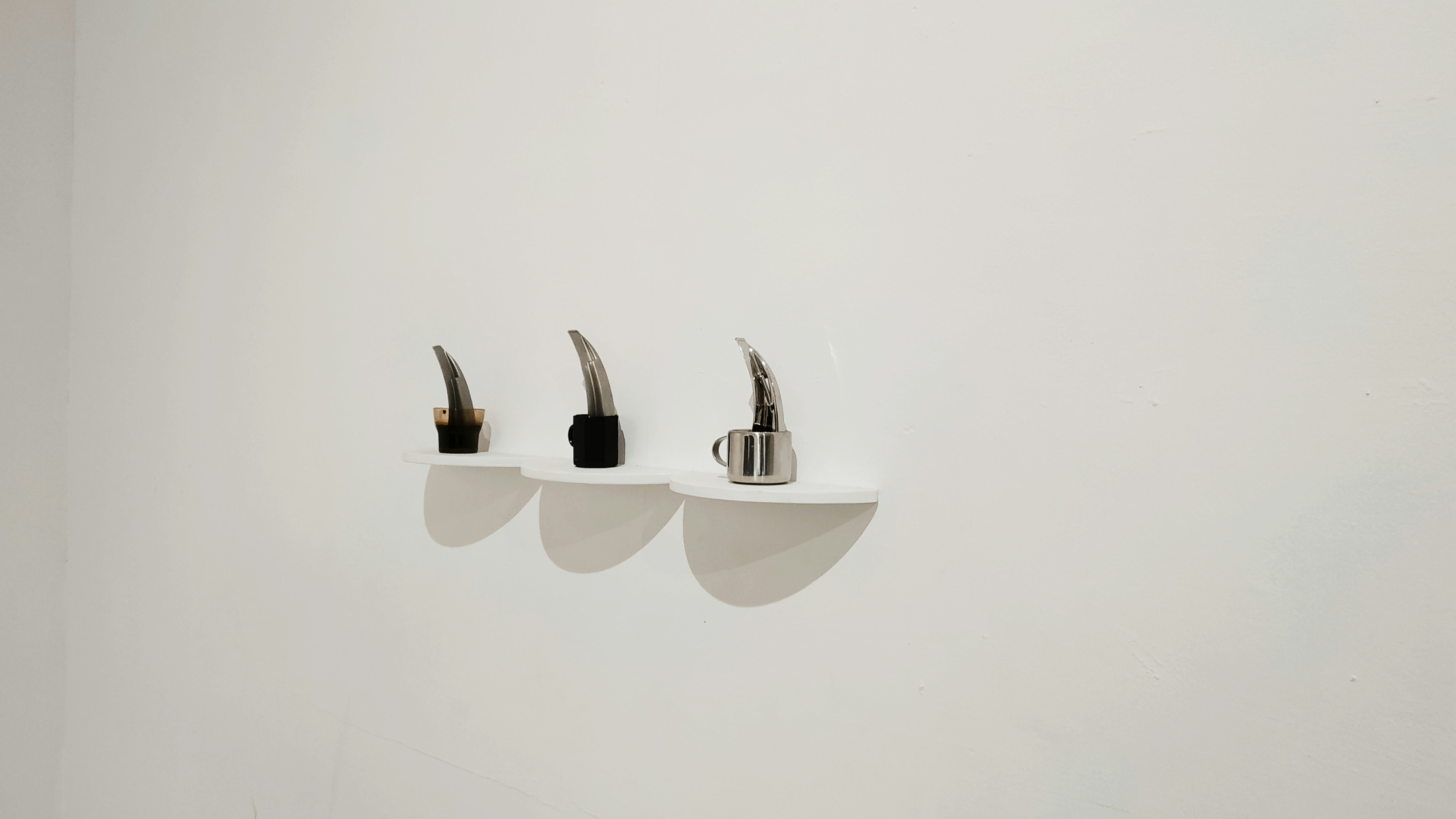 Three uniquely shaped metallic objects arranged on a minimalist shelf against a stark white wall.