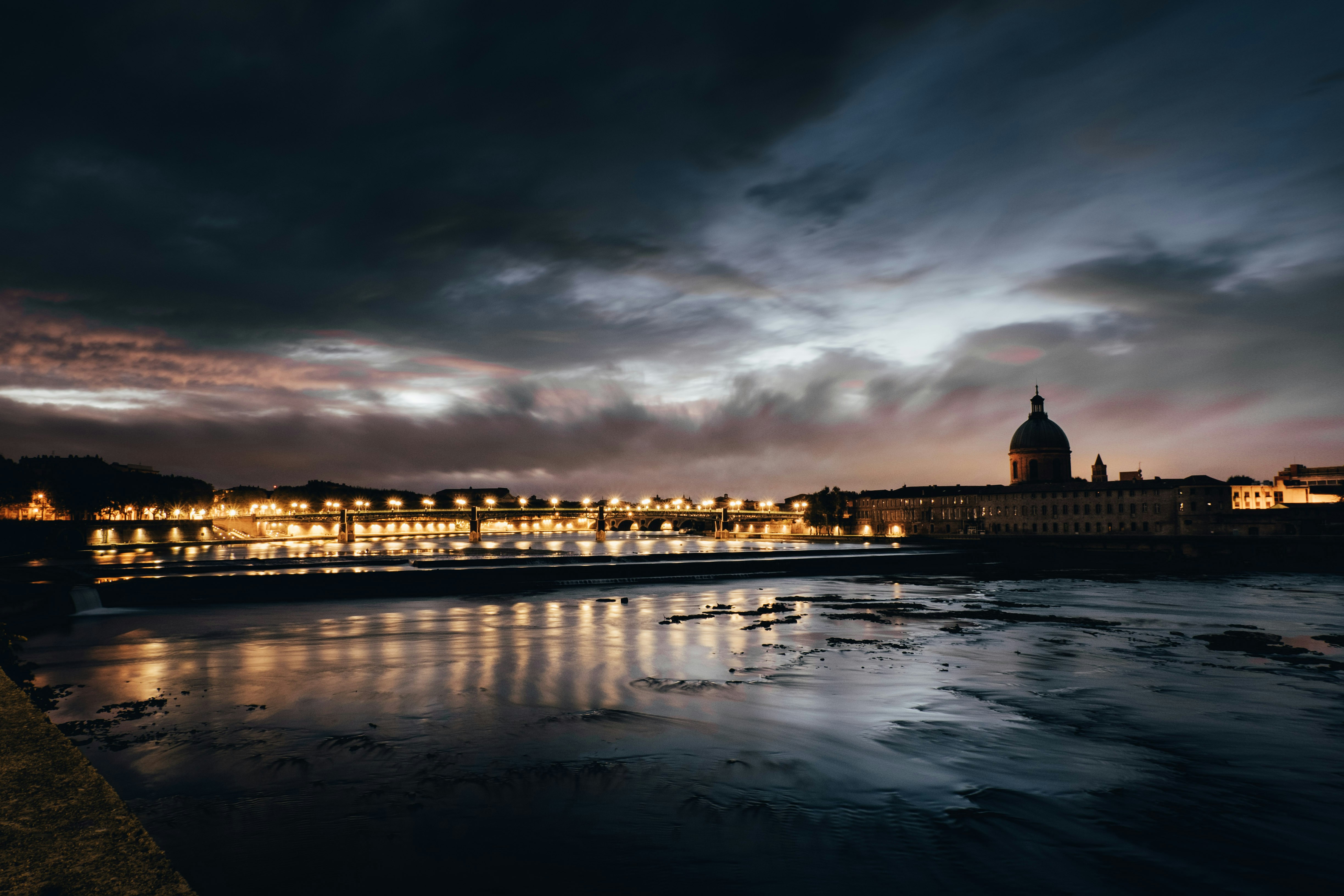 Illuminated bridge and dome against a dramatic twilight sky reflecting on the river in Toulouse.