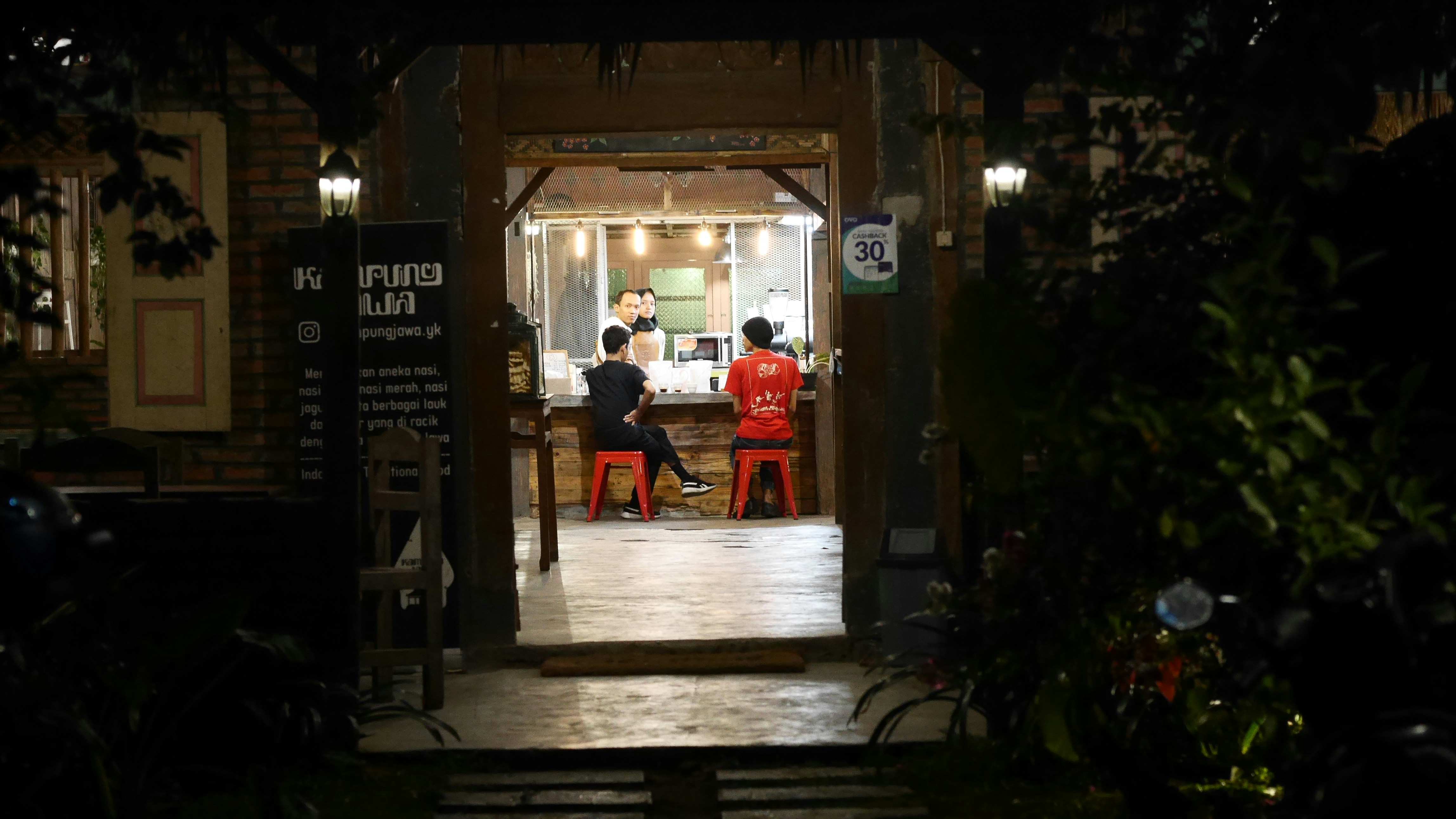 Two people sit on red stools at a counter inside a warmly lit café, viewed from an open doorway surrounded by foliage.