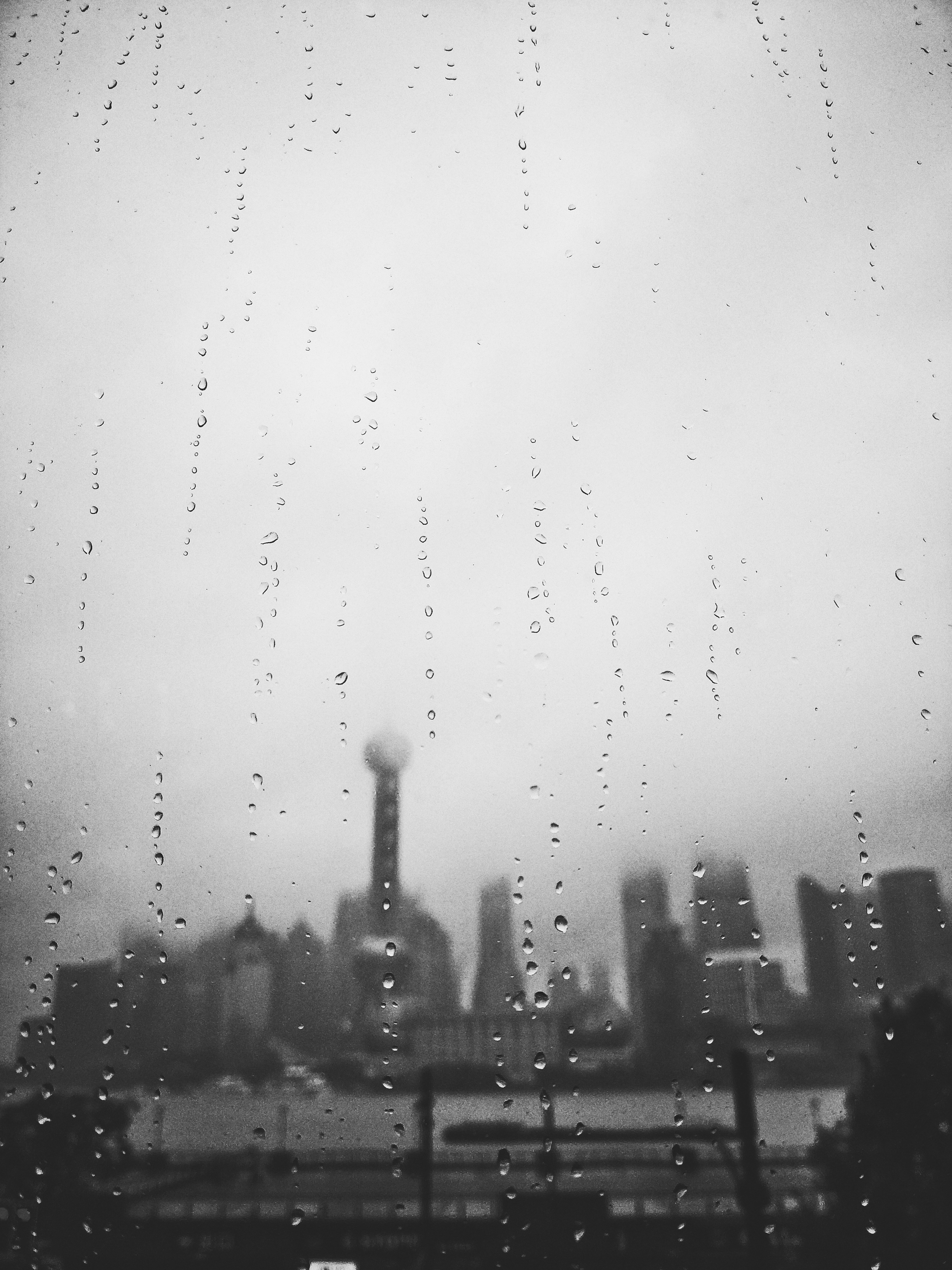 Raindrops cascade down a window, obscuring a silhouette of a city skyline under a gray sky.