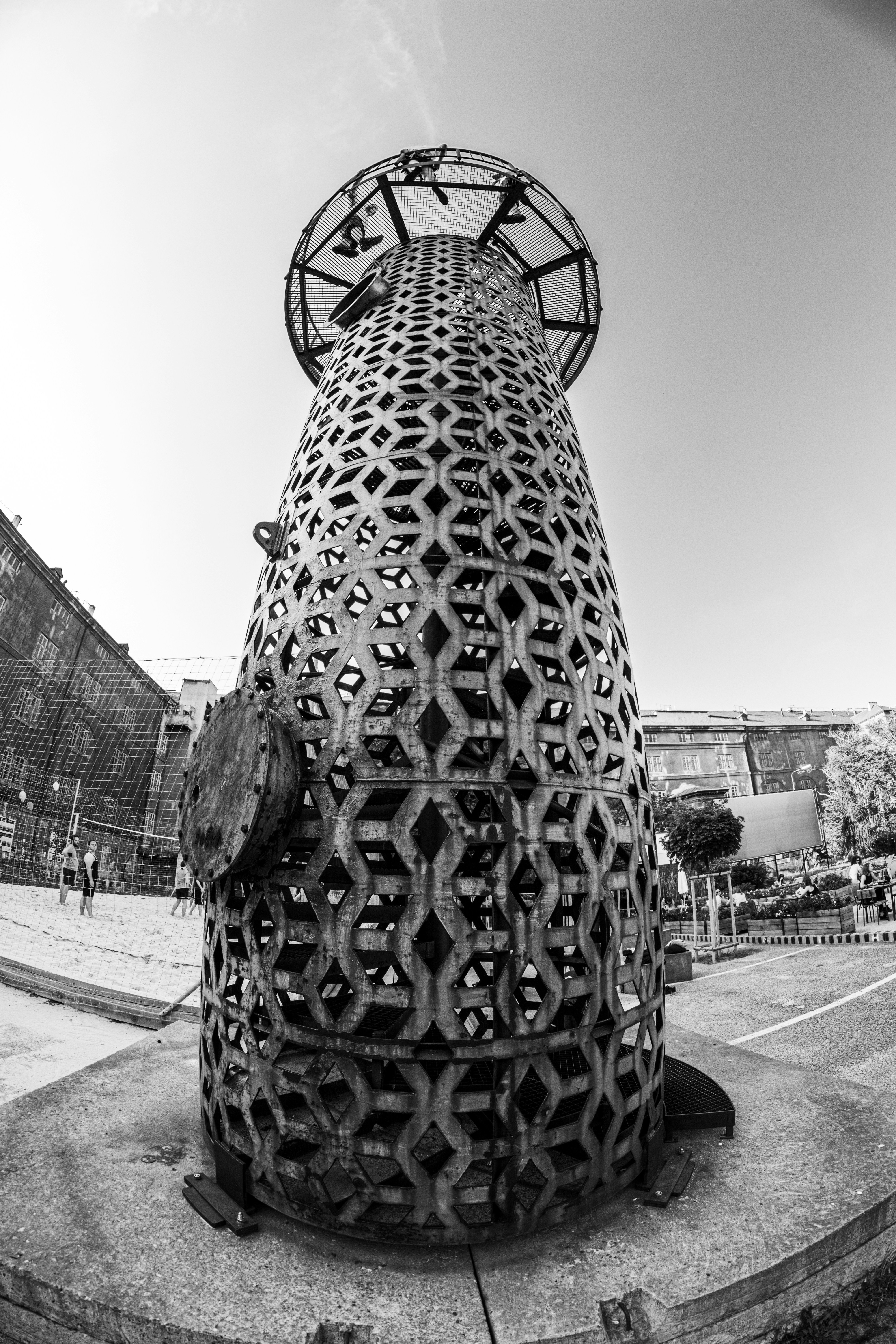 Intricate metal tower with geometric patterns stands prominently in an urban park setting, surrounded by greenery and activity.