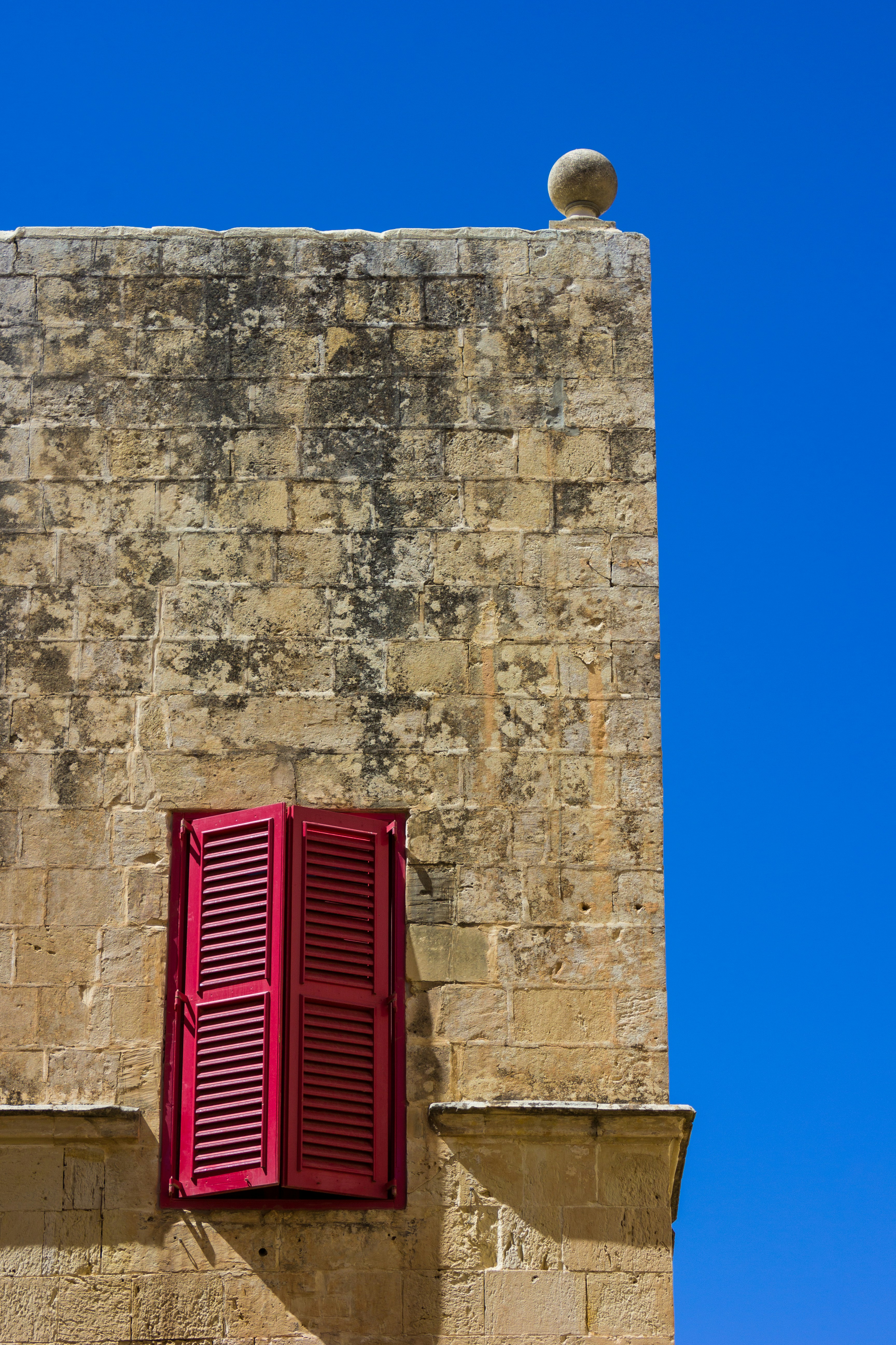 Bright red shutters against a weathered stone wall under a clear blue sky highlight the charm of traditional architecture.