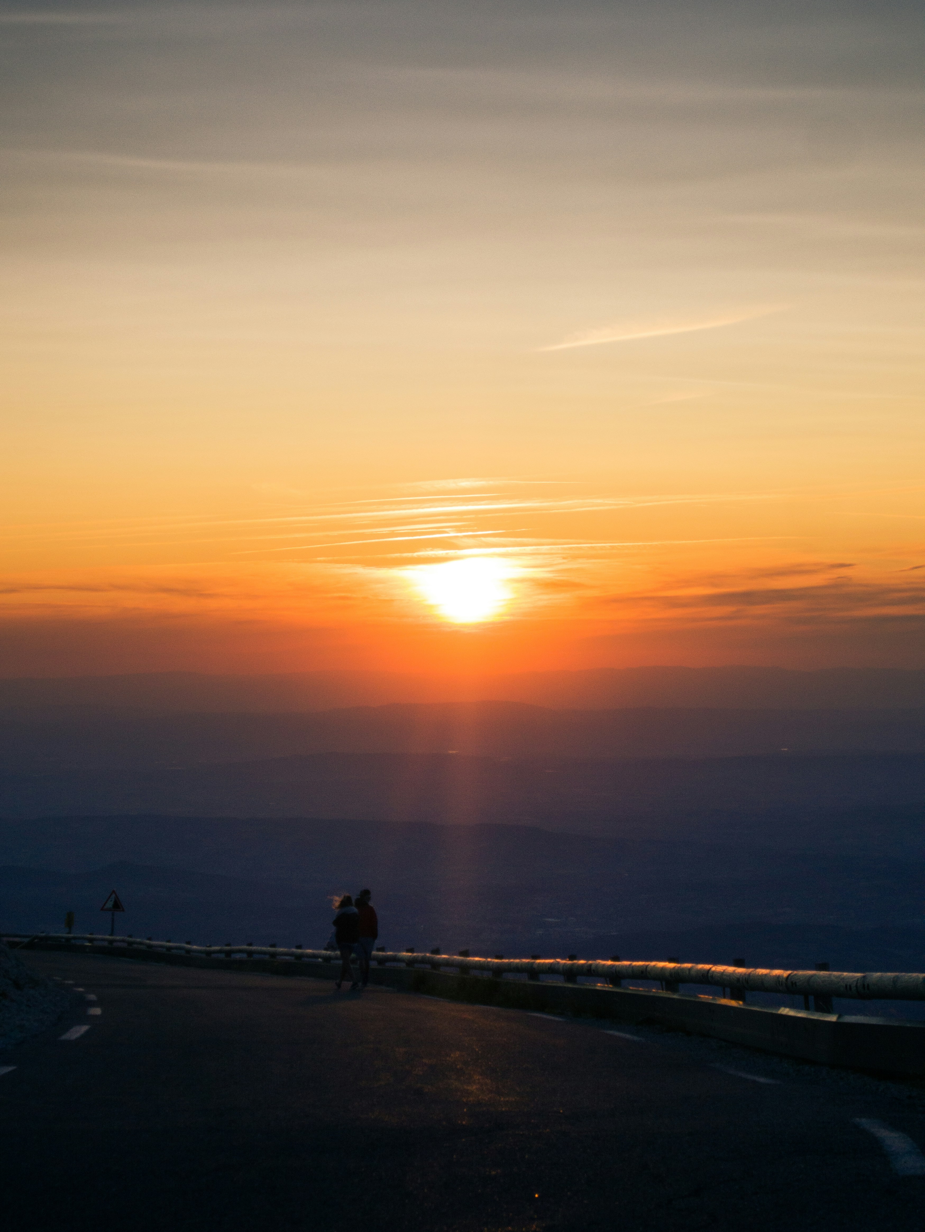 Silhouetted couple walking along a winding road as the sun sets over distant mountains, casting warm hues across the sky.