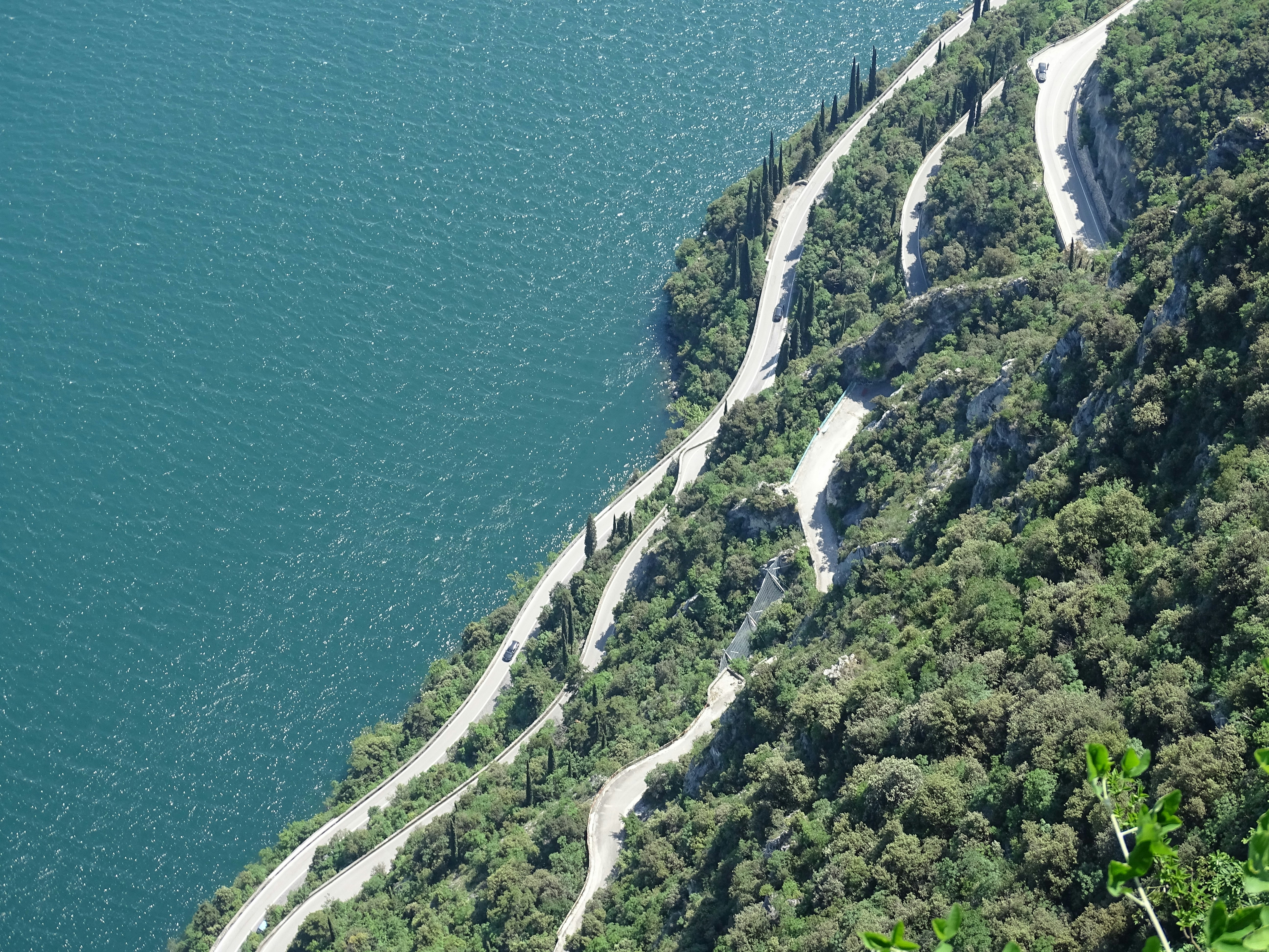 Aerial view of winding roads cutting through lush greenery beside a deep blue body of water.