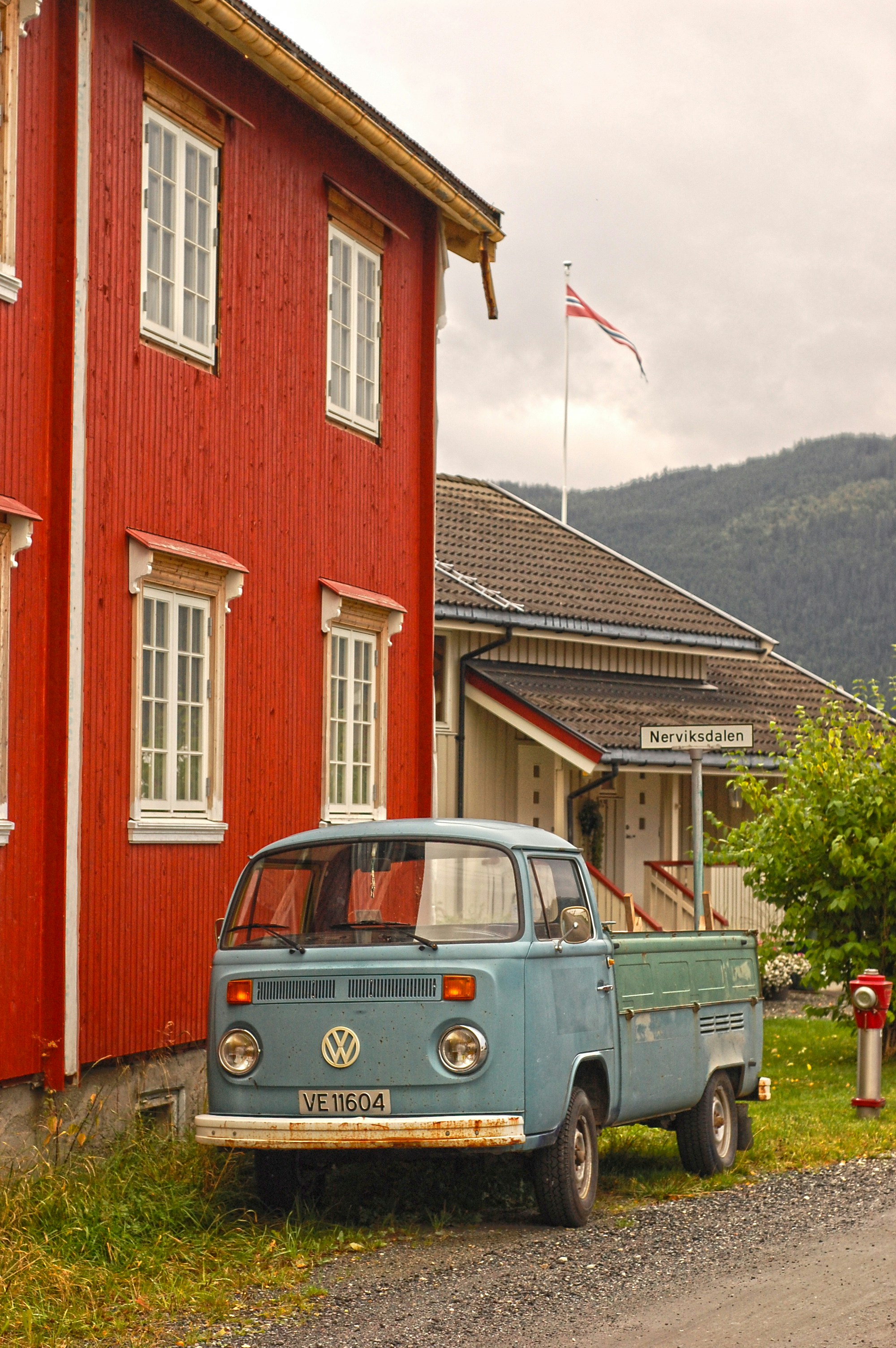 Classic blue Volkswagen truck parked beside a vibrant red wooden house, with a backdrop of lush hills and a flag fluttering in the breeze.