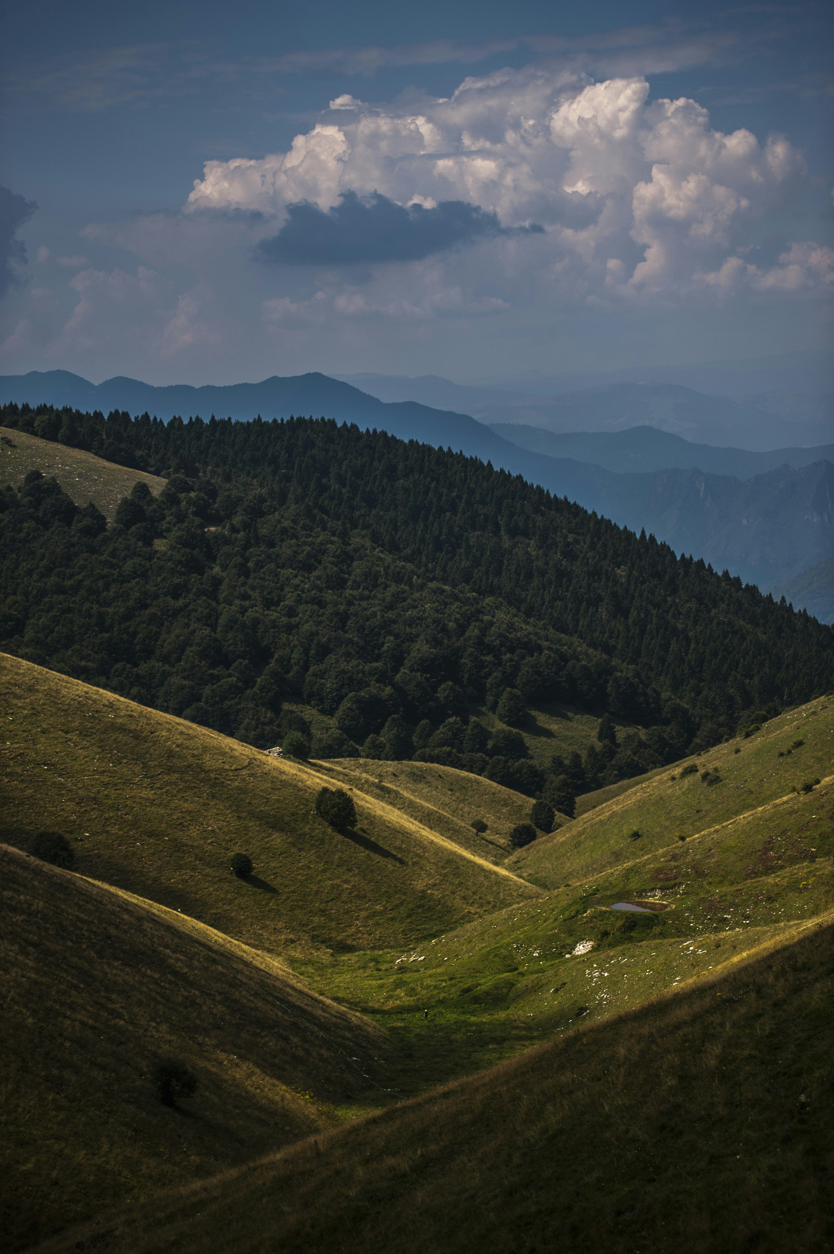 Gentle hills adorned with patches of greenery and sparse trees, set against a backdrop of distant mountains and clouds. The landscape captures the serene beauty of nature.
