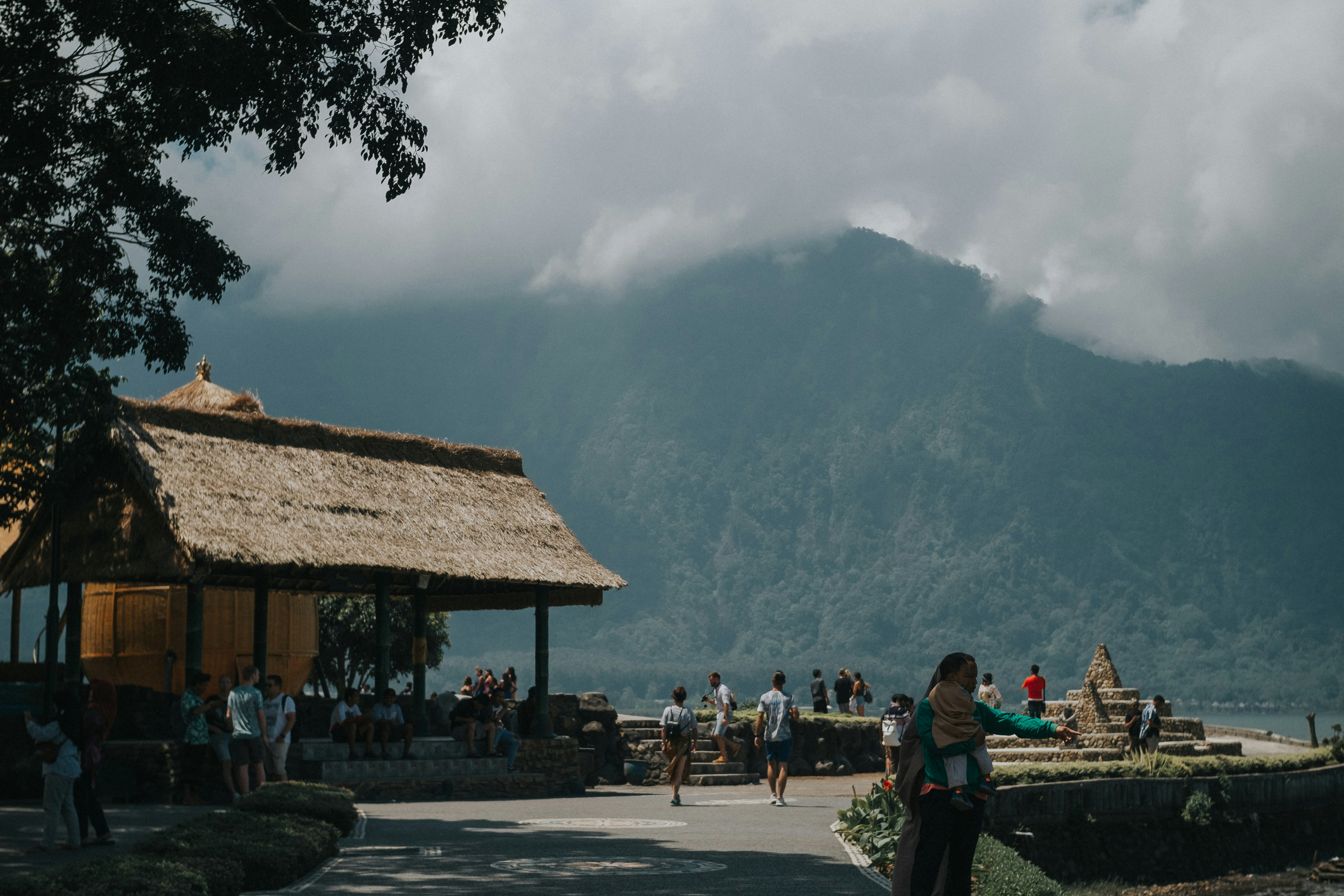 Lakeside pavilion with a thatched roof along a promenade, groups of visitors by the water, with cloud-covered mountains in the background.