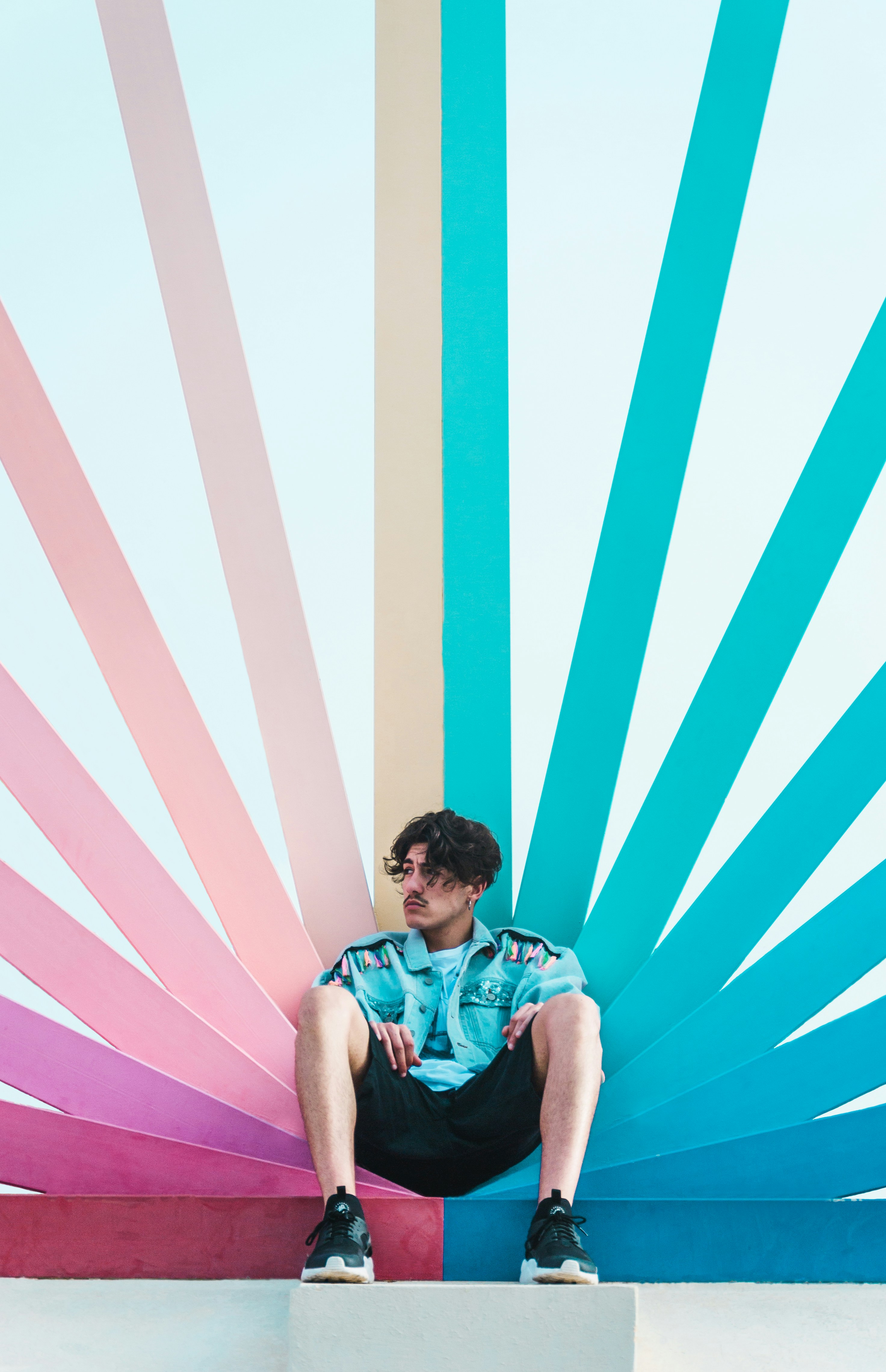 A young man seated against a vibrant backdrop of pastel rays, showcasing a blend of pinks, blues, and yellows. His relaxed pose contrasts with the dynamic lines of the mural.