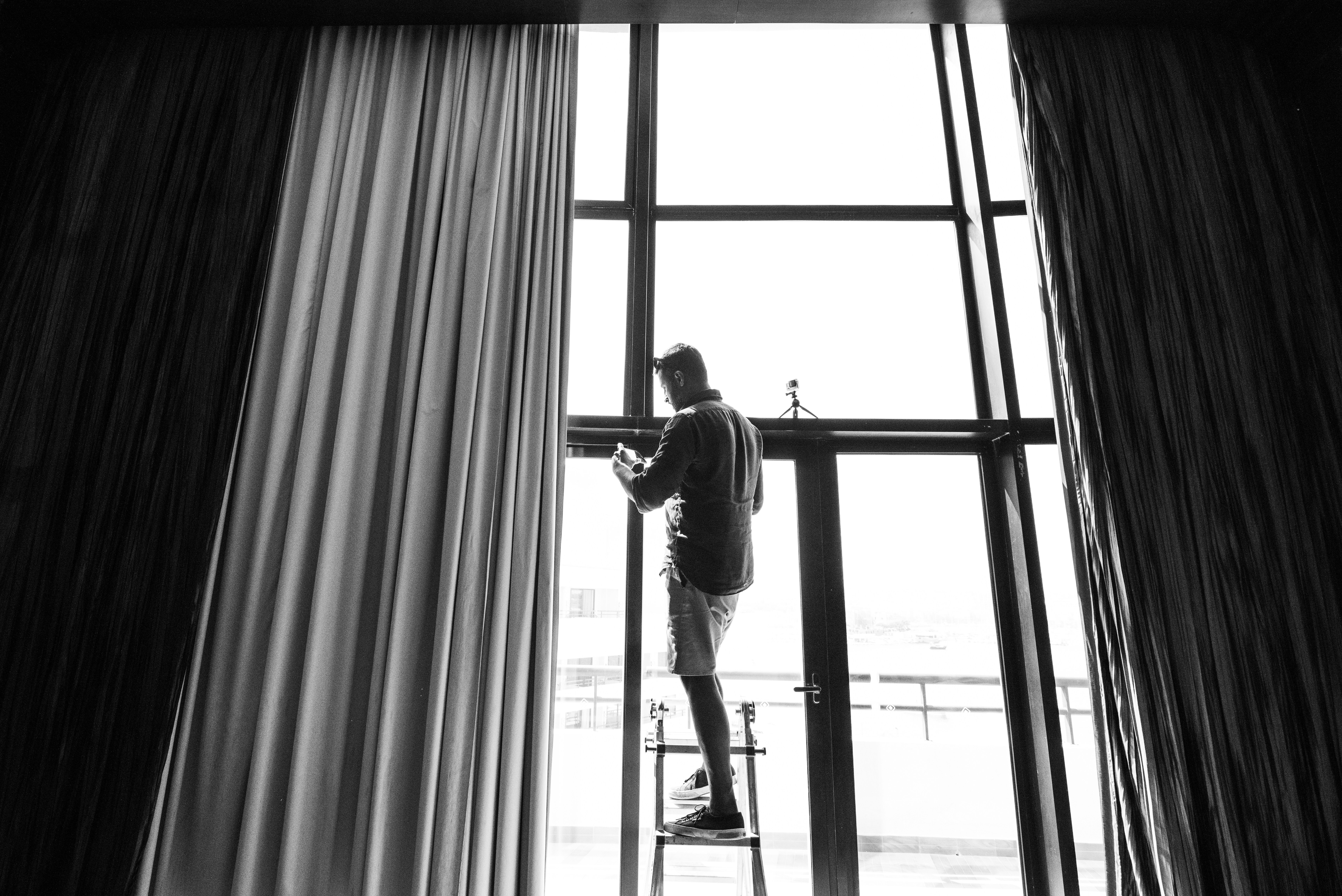 A worker adjusts curtains while standing on a ladder in front of large windows, framed by flowing drapes. The scene is set in a modern interior space.