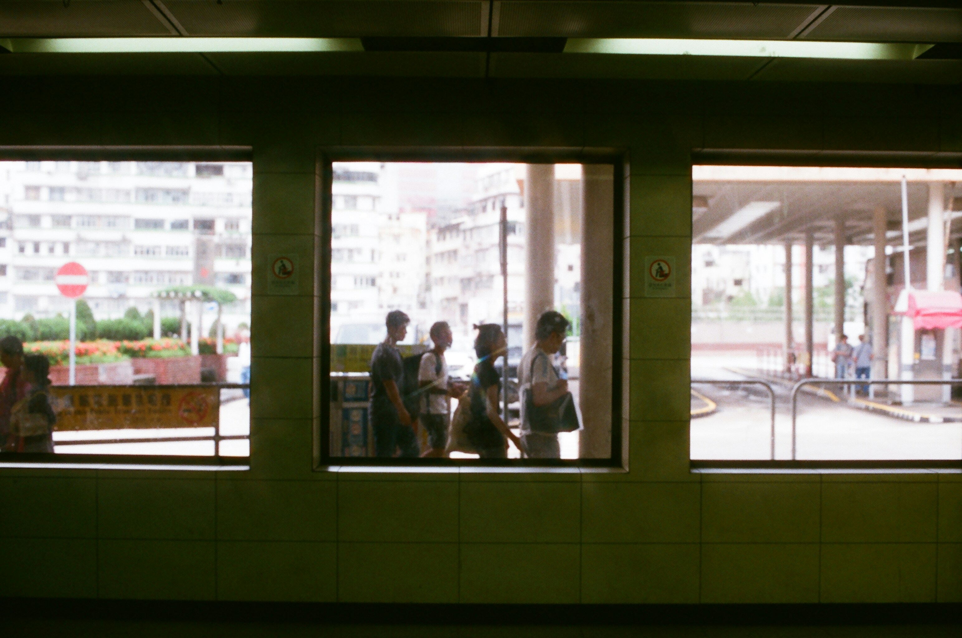 Silhouetted figures moving through a transit station, framed by large windows that reveal a bustling urban backdrop.