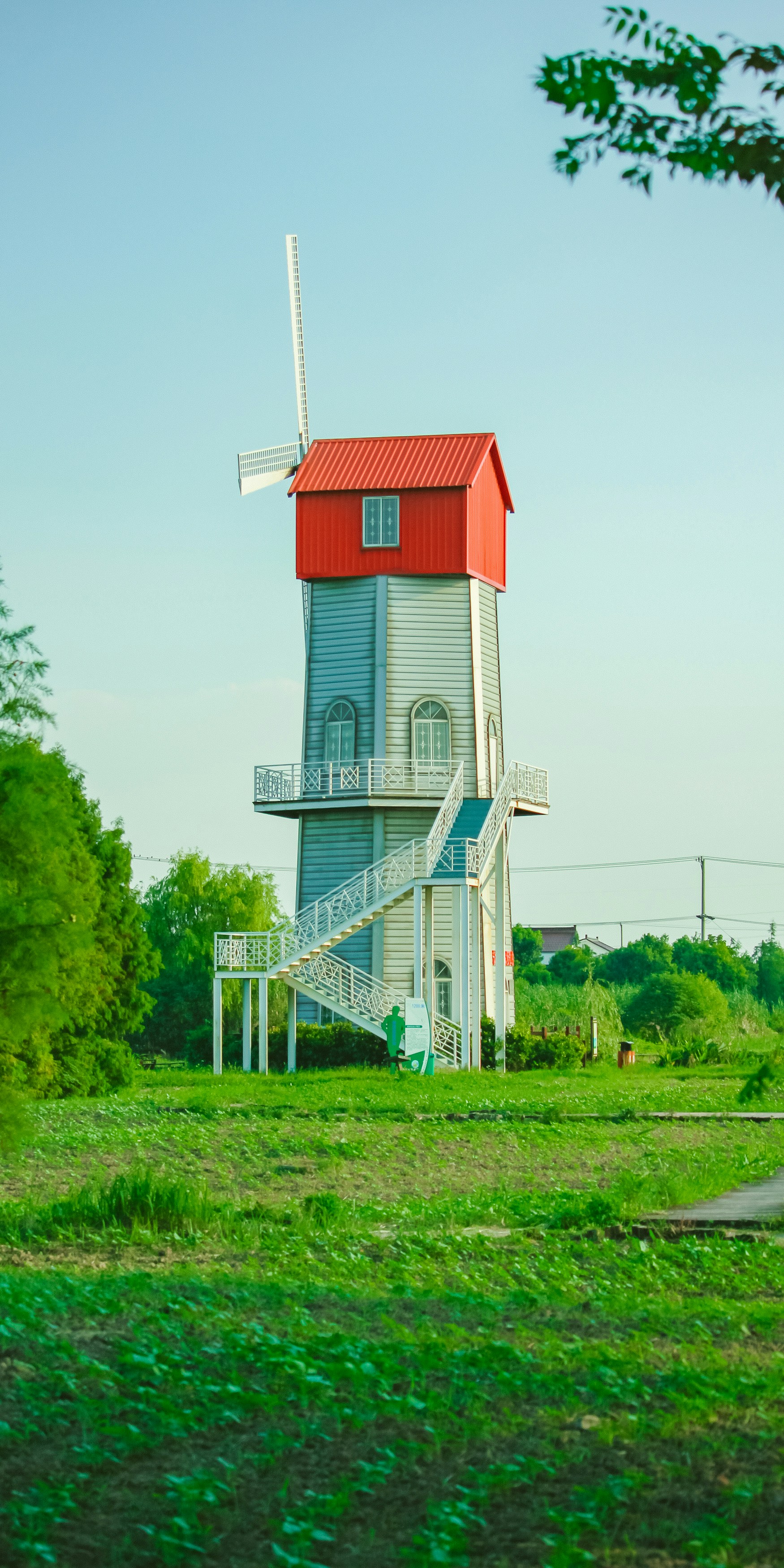 A charming windmill with a vibrant red roof stands against a clear sky, surrounded by lush greenery and a well-kept lawn.