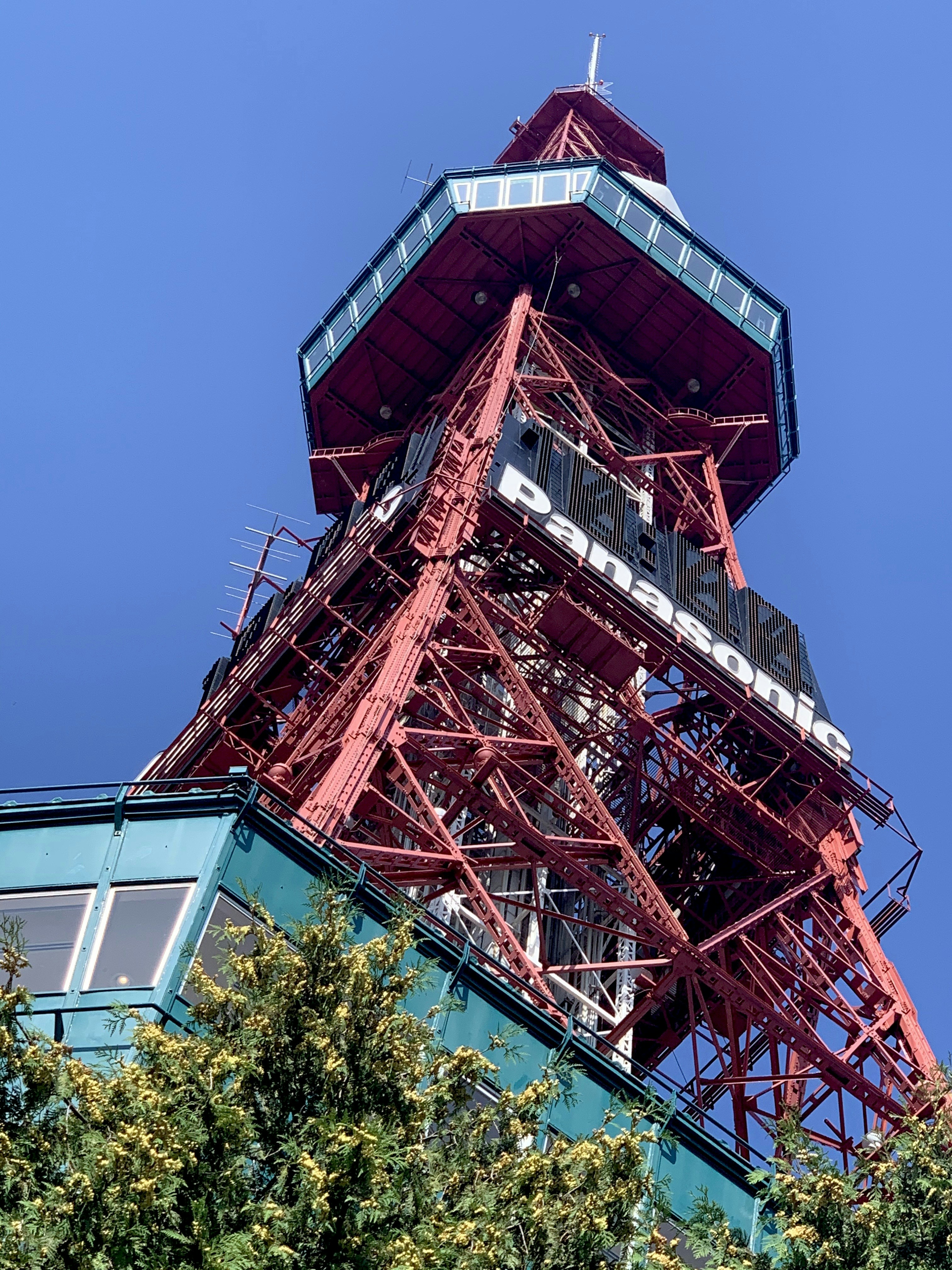 Red steel broadcasting tower with 'Panasonic' signage against a clear blue sky, surrounded by green foliage.