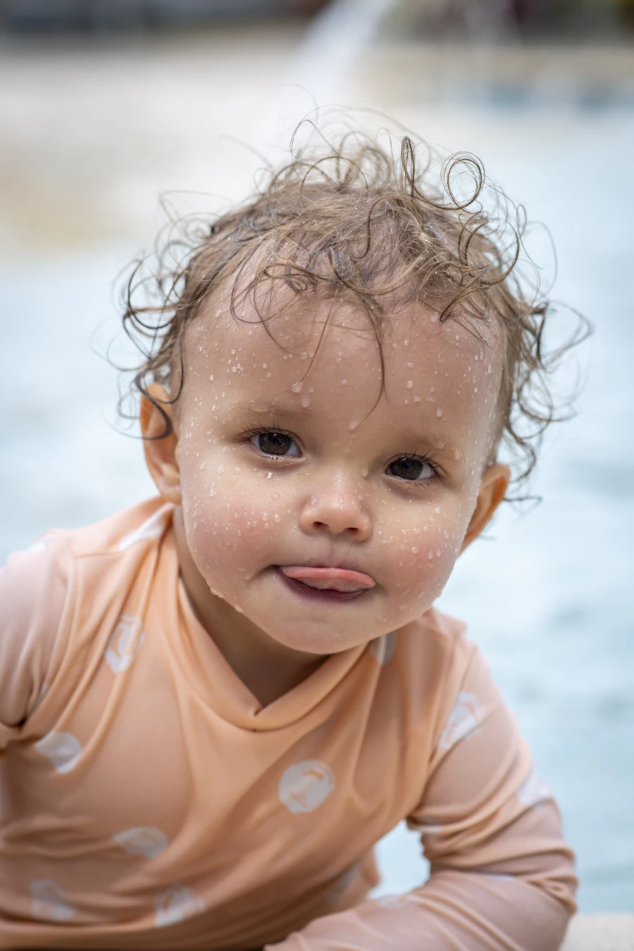 Brightly lit portrait of a wet toddler by a pool, cheeks rosy, tongue playfully peeking out, peach swimsuit, water backdrop. A candid close-up emphasizing playful expression.