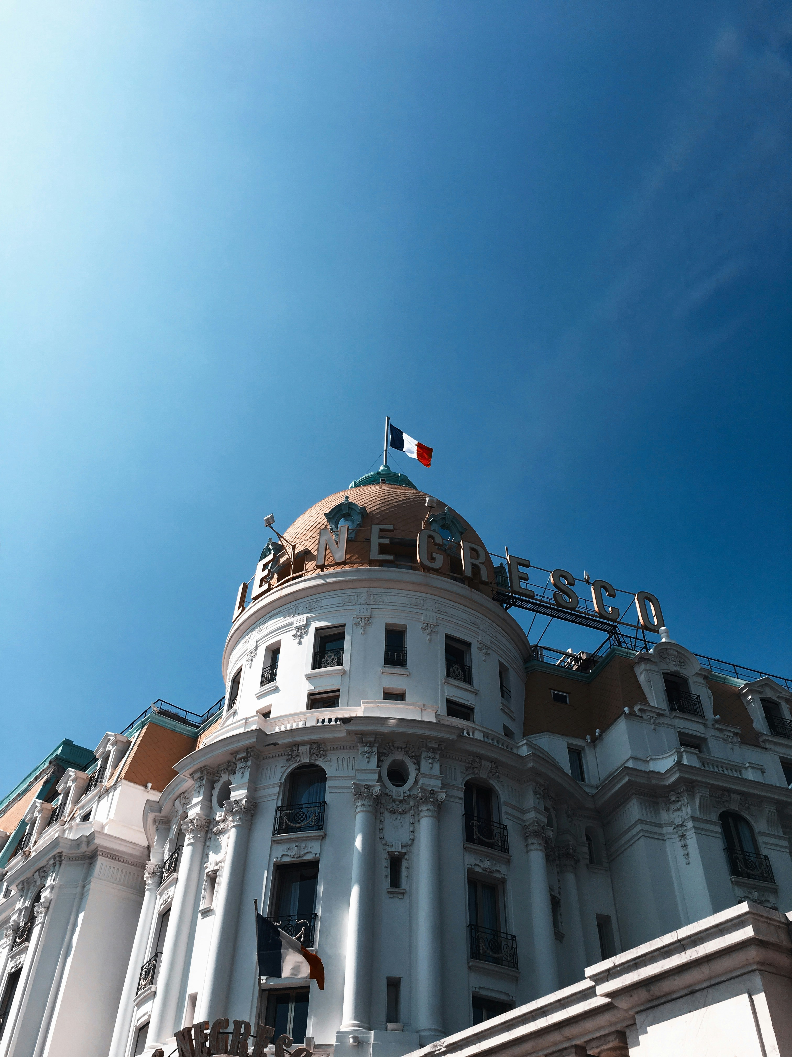 Historic hotel facade adorned with a French flag against a clear blue sky. Architectural details highlight its grandeur.