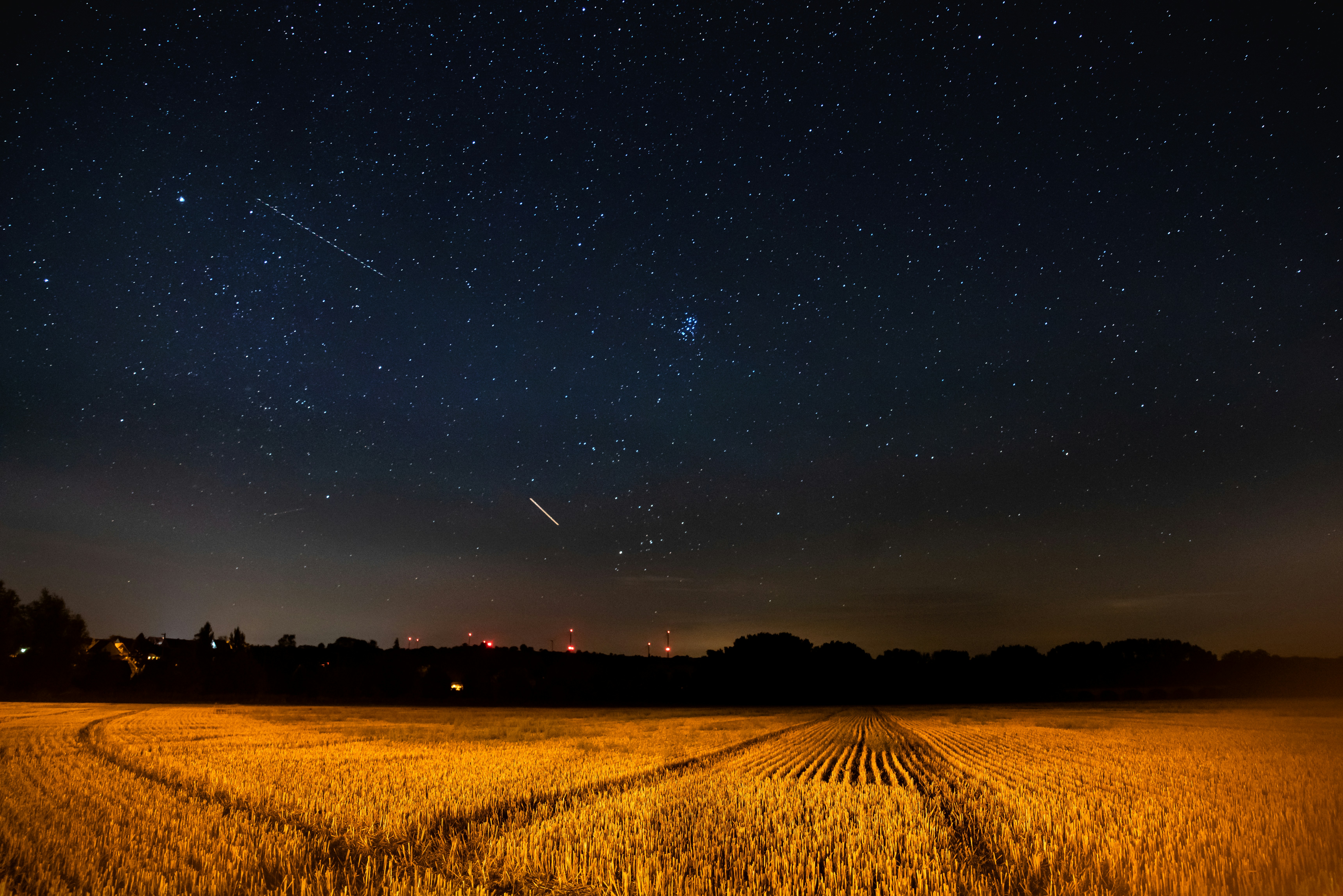 Vast starry night sky over a golden wheat field, with faint traces of shooting stars and distant wind turbines illuminating the horizon.