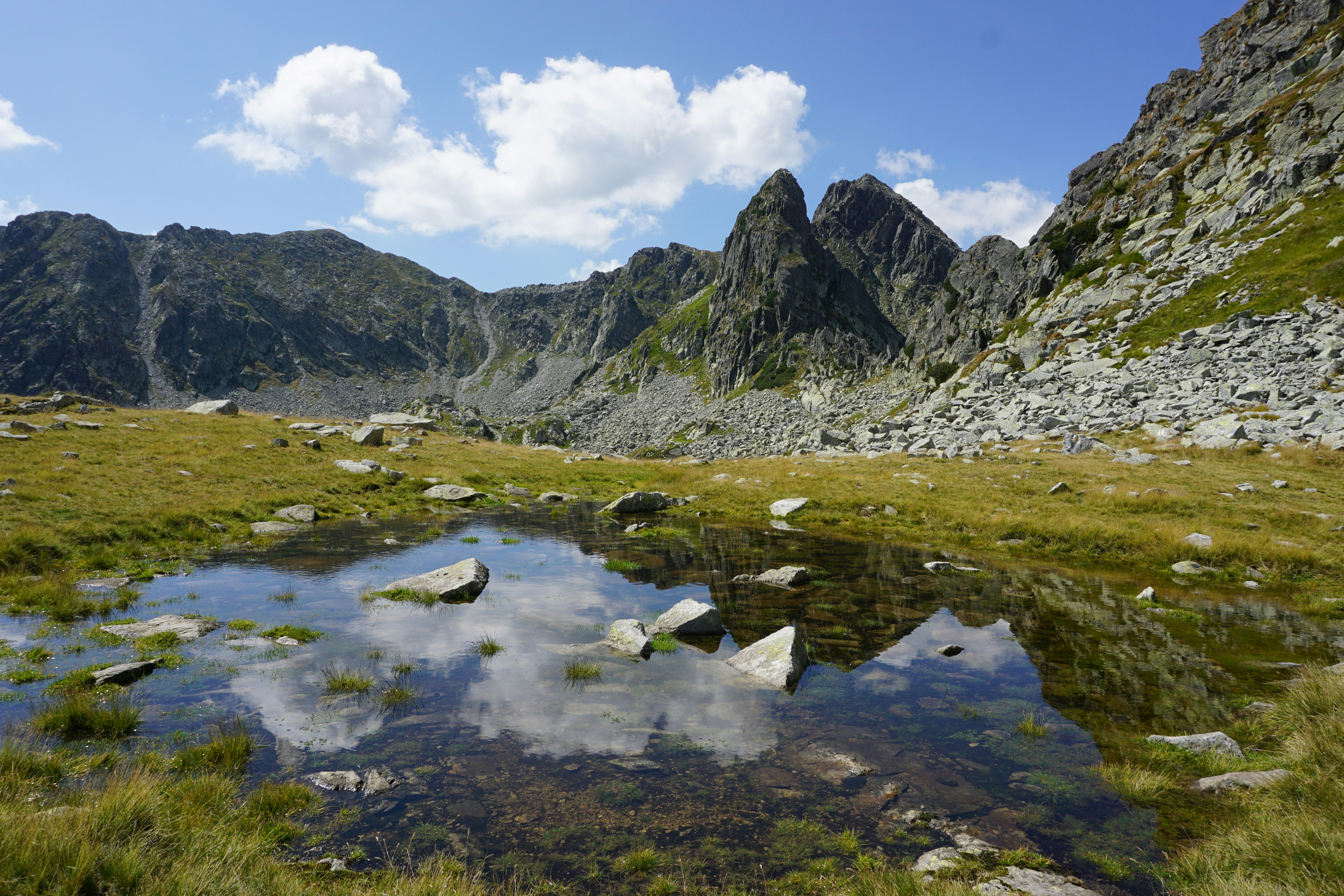 Rocky peaks reflected in a tranquil mountain pond under a bright blue sky.