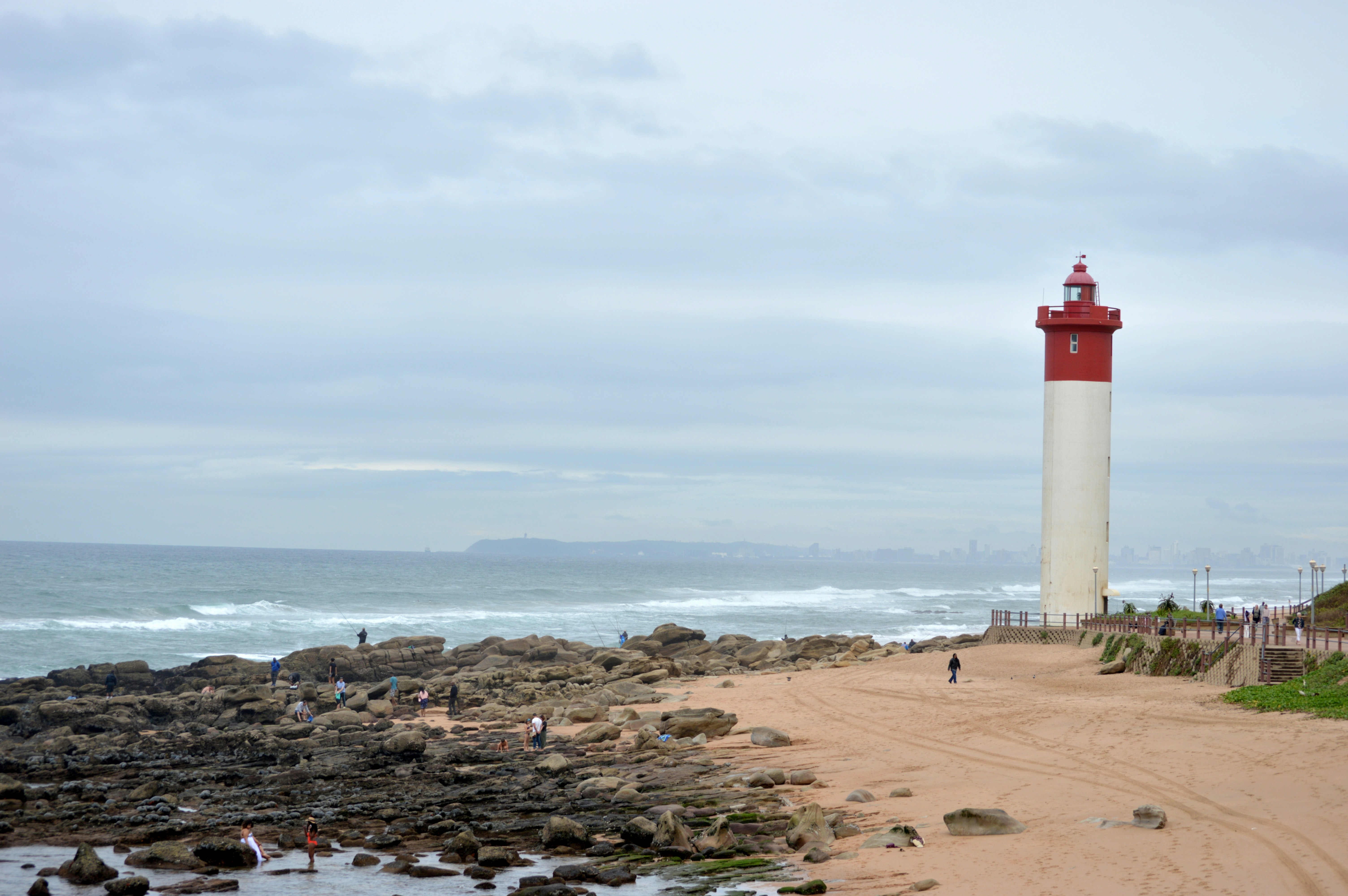 White and red lighthouse stands near rocky shore with ocean waves under a cloudy sky.
