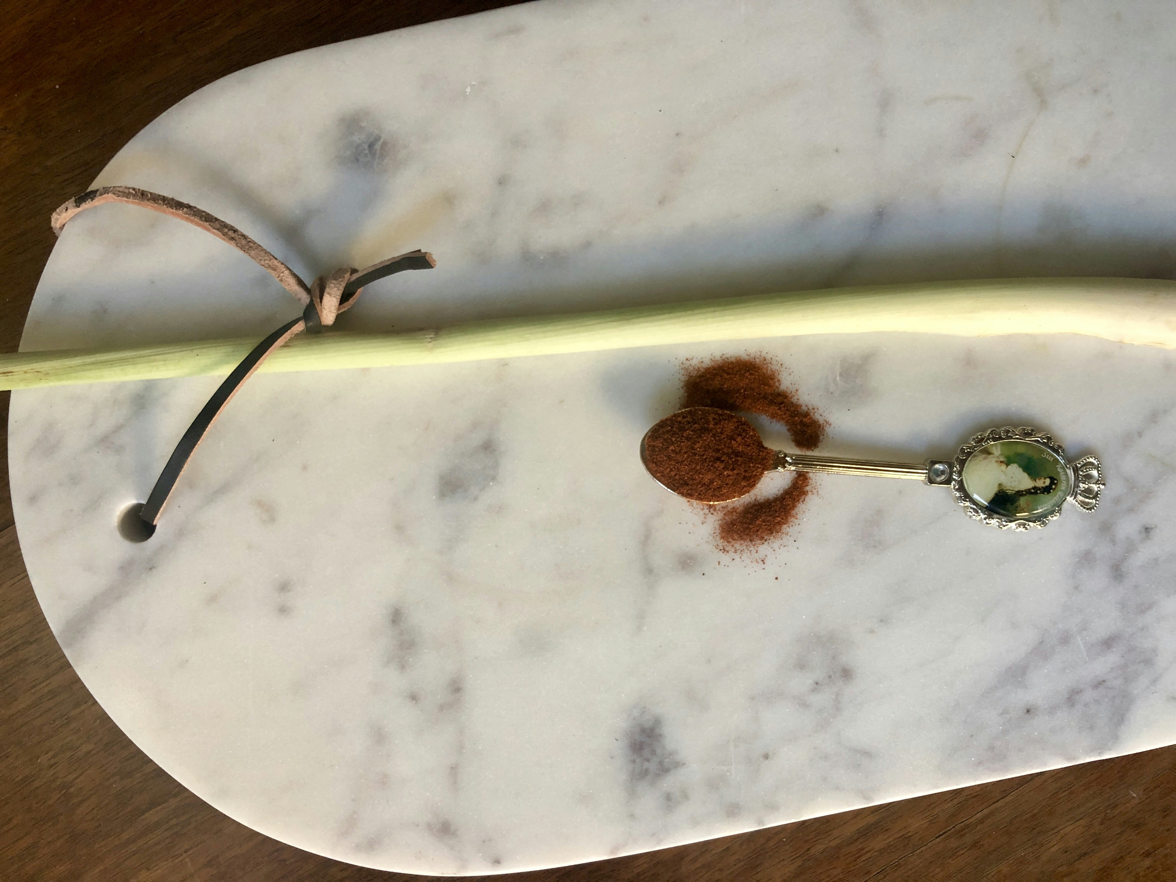 A vintage spoon adorned with a delicate design rests beside a sprinkle of spice on a marble cutting board, complemented by a green onion stalk.