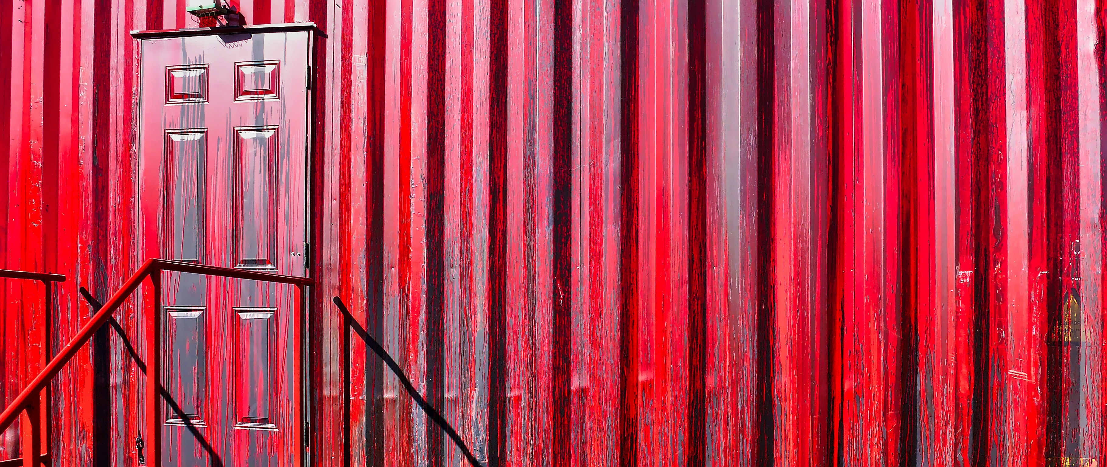 Red corrugated metal wall with a gray door on the left and a small bird perched near the top center.