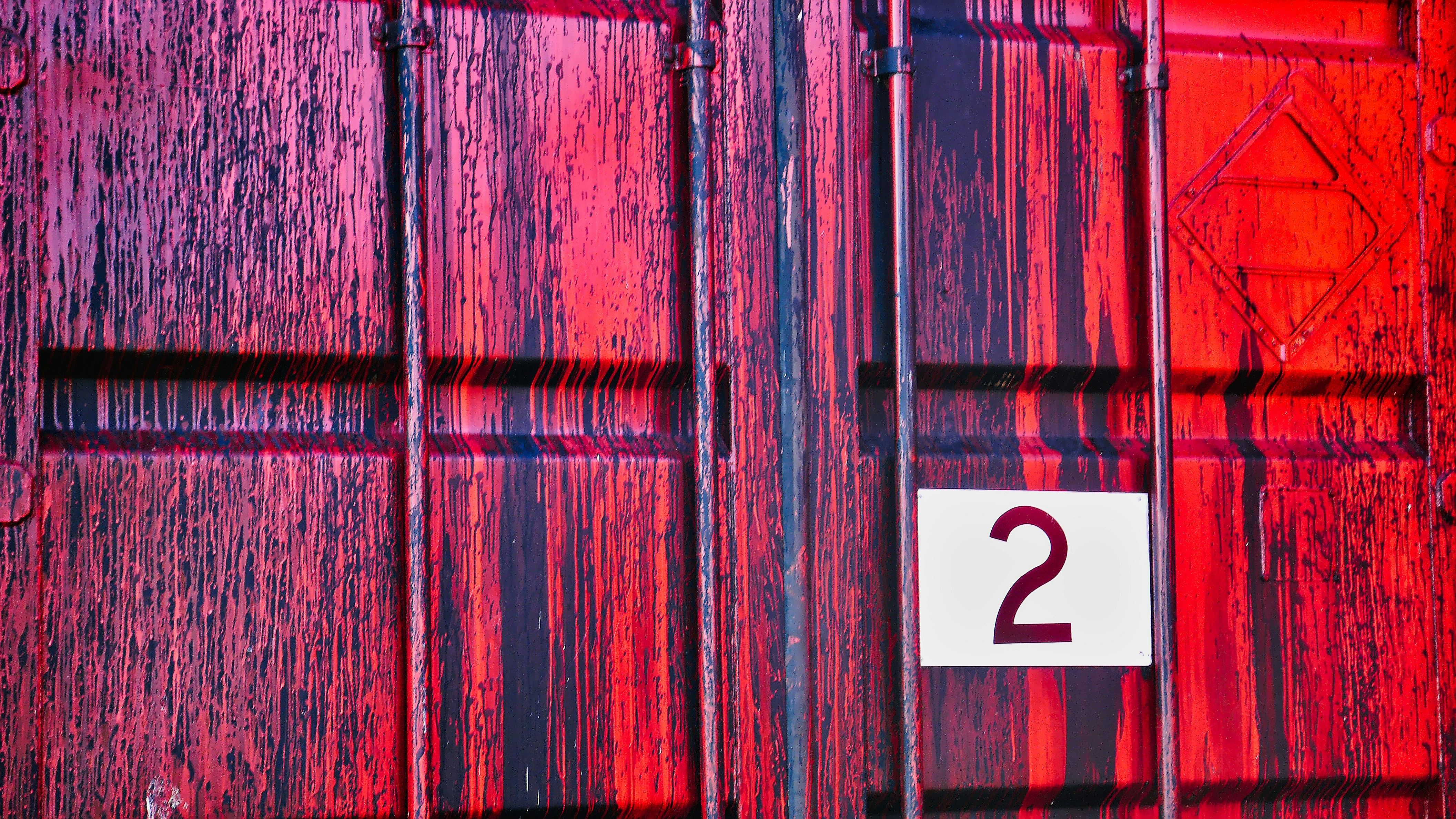 Close-up photograph of weathered red doors with peeling paint and a white numbered plaque displaying the number 2.