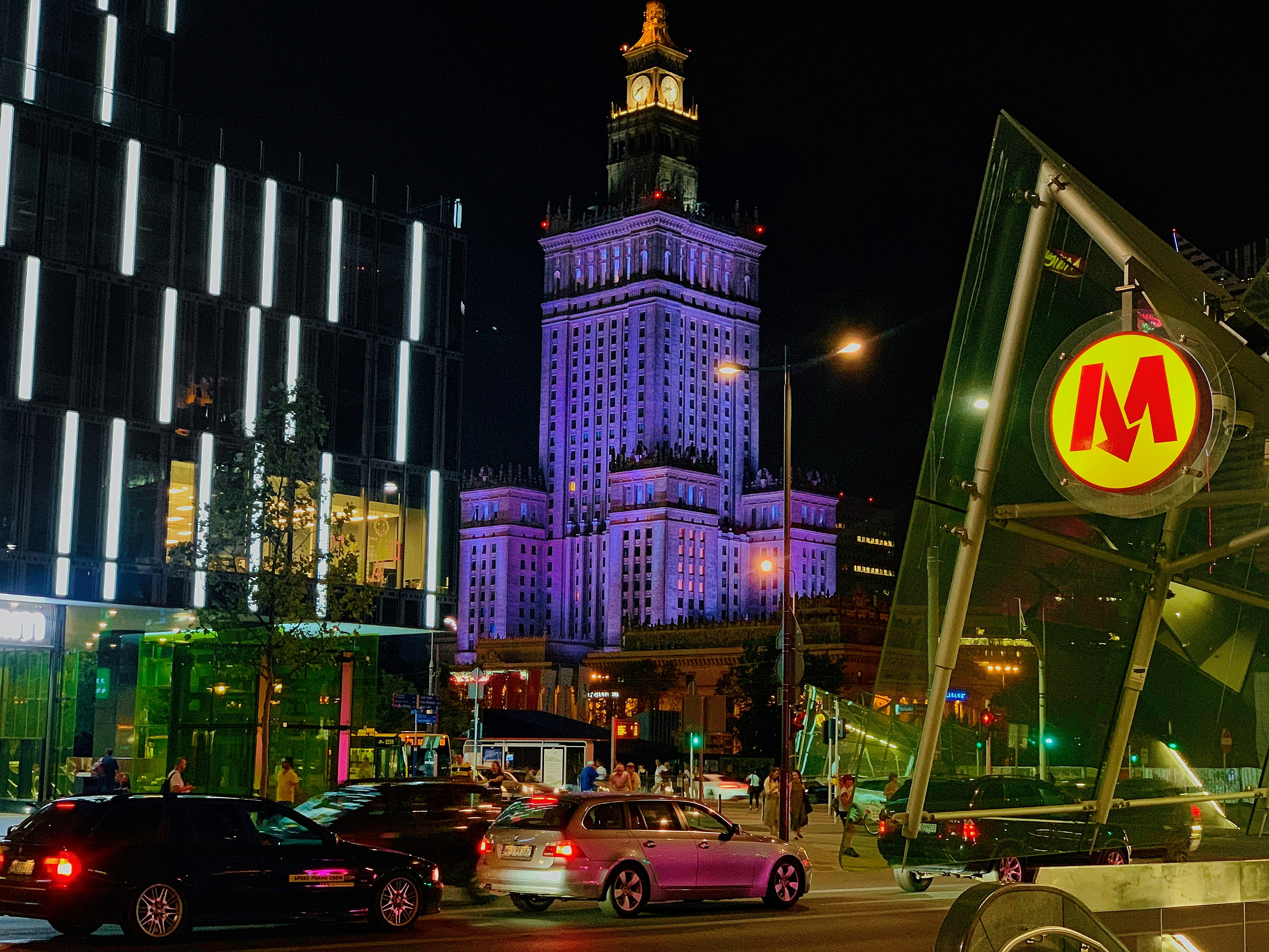 Illuminated skyscraper towers over bustling street traffic at night, with a vibrant subway logo in the foreground.
