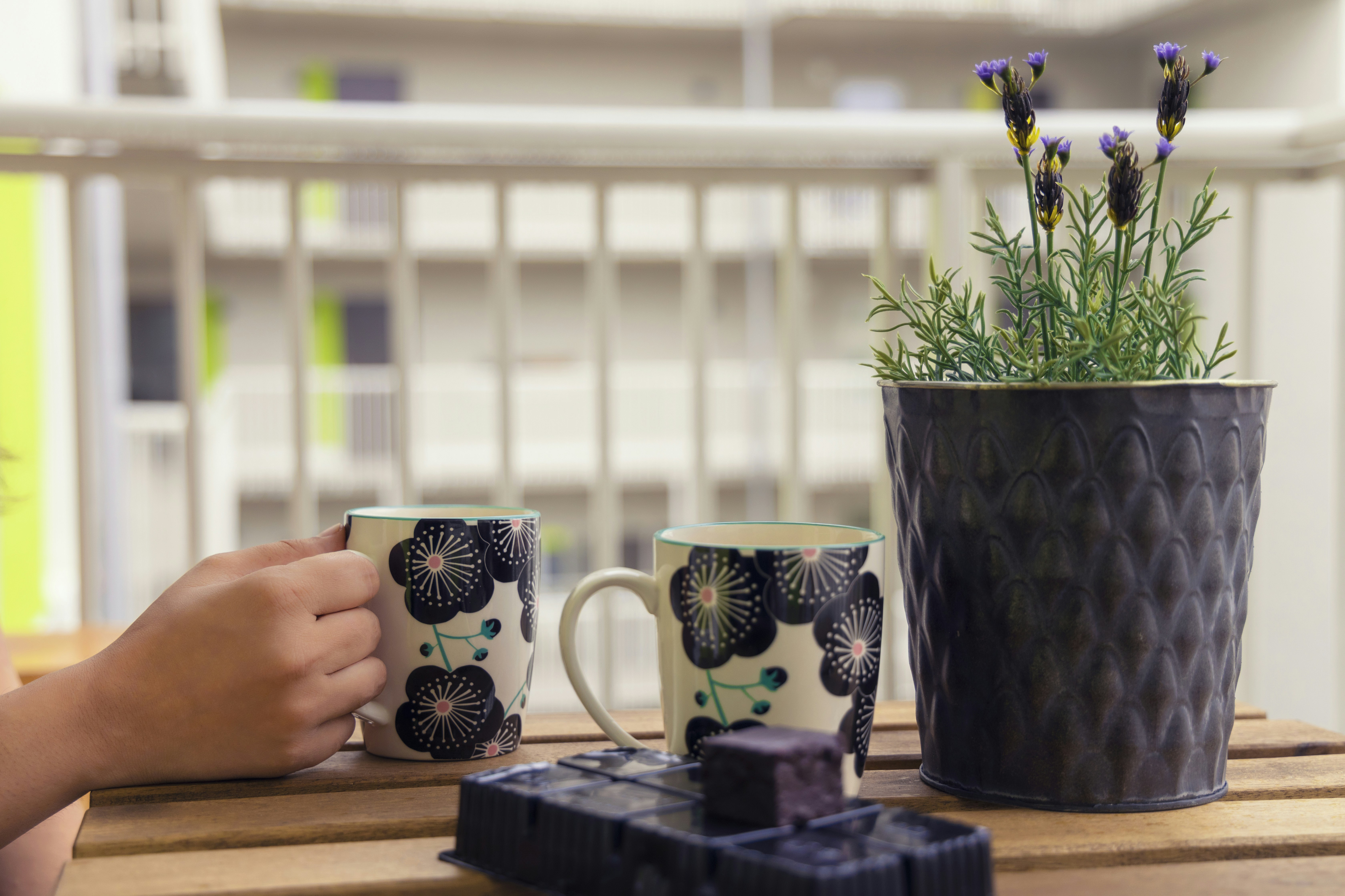 Hand holding a floral mug near a potted lavender plant on a wooden balcony table.