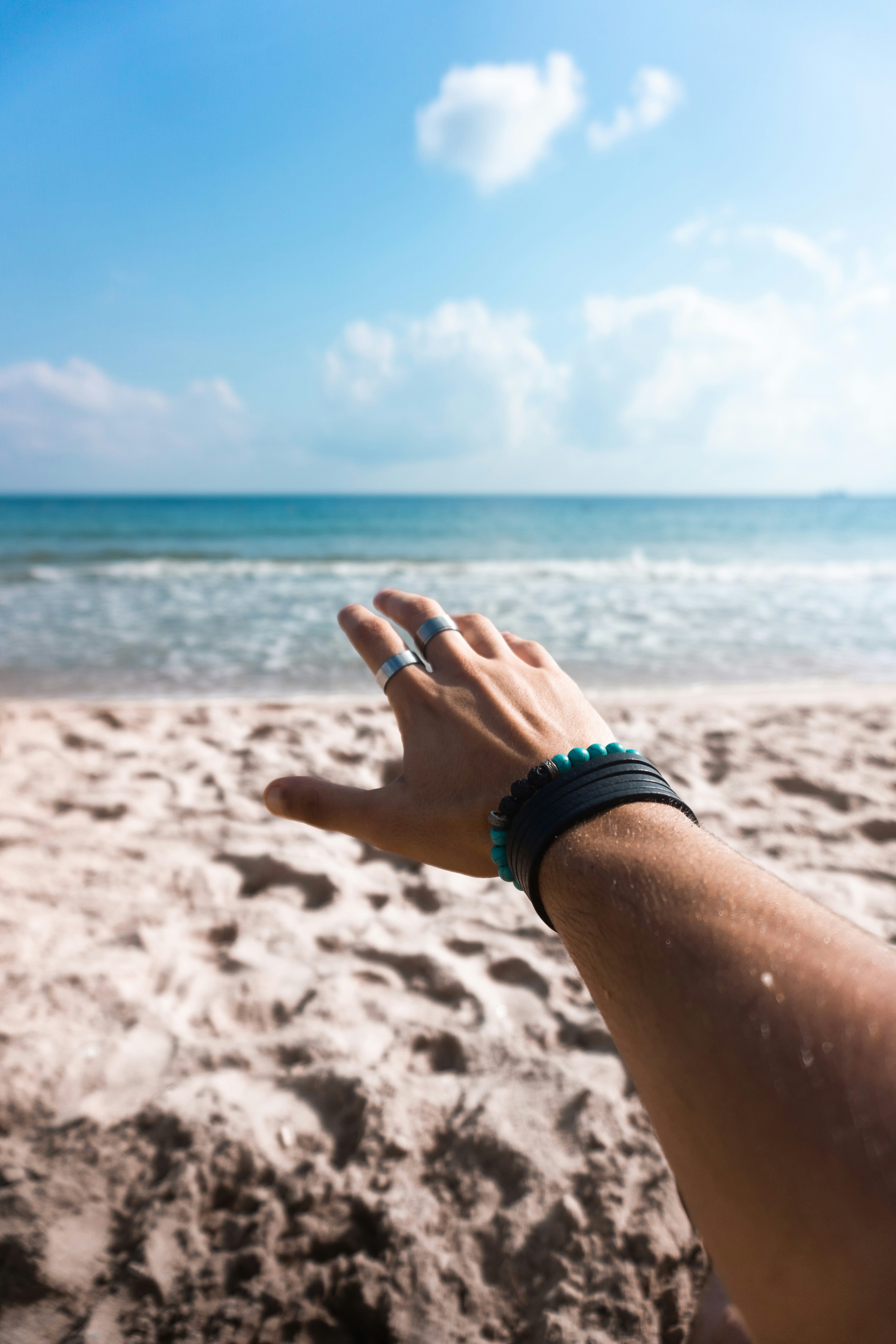 A hand adorned with bracelets extends toward the tranquil ocean, capturing a moment of connection with nature. The soft sand and gentle waves create a serene beach atmosphere.