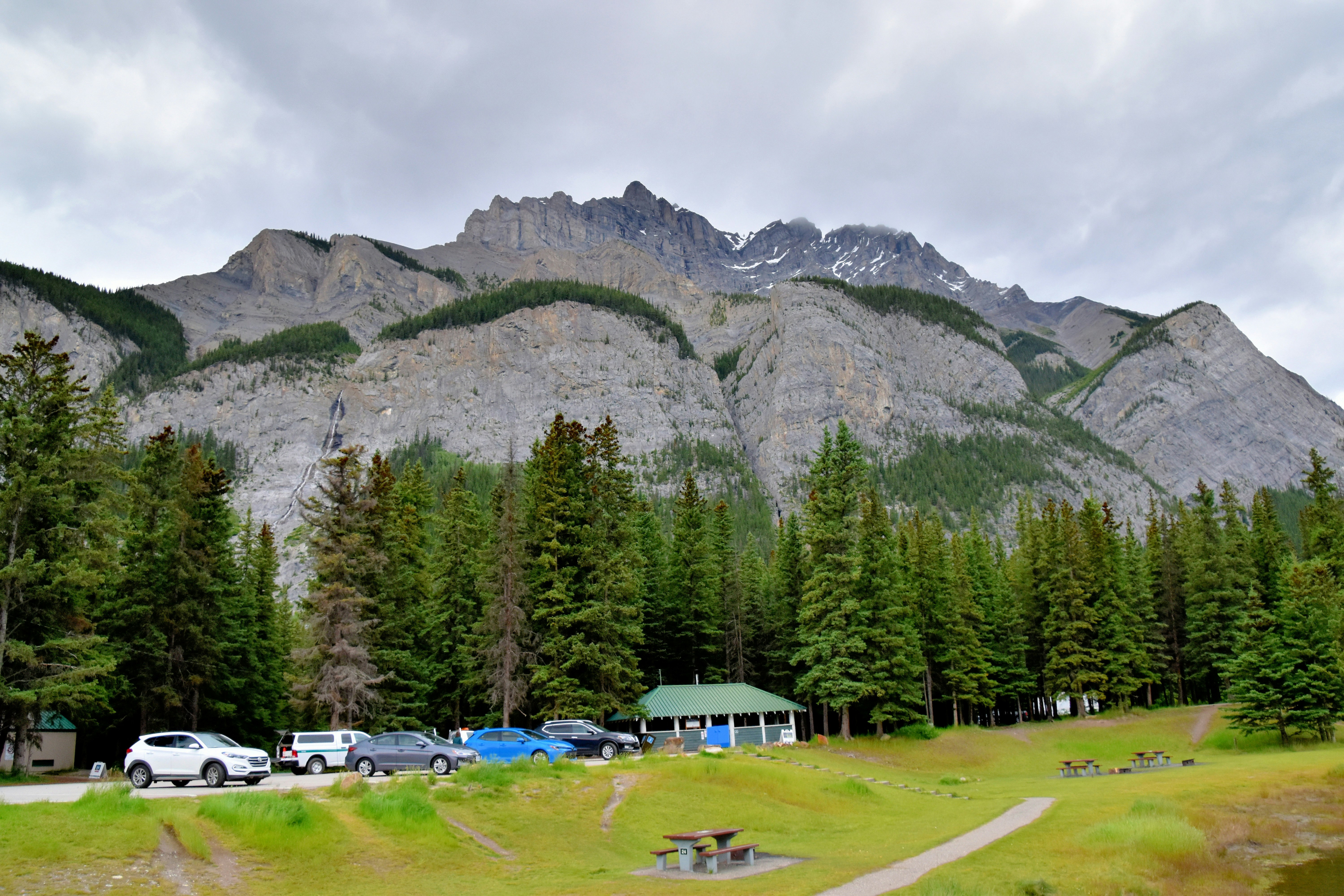 A wide mountain face rises behind a line of evergreen trees, with a campground and parked cars at the grassy foreground, conveying a tranquil wilderness scene.
