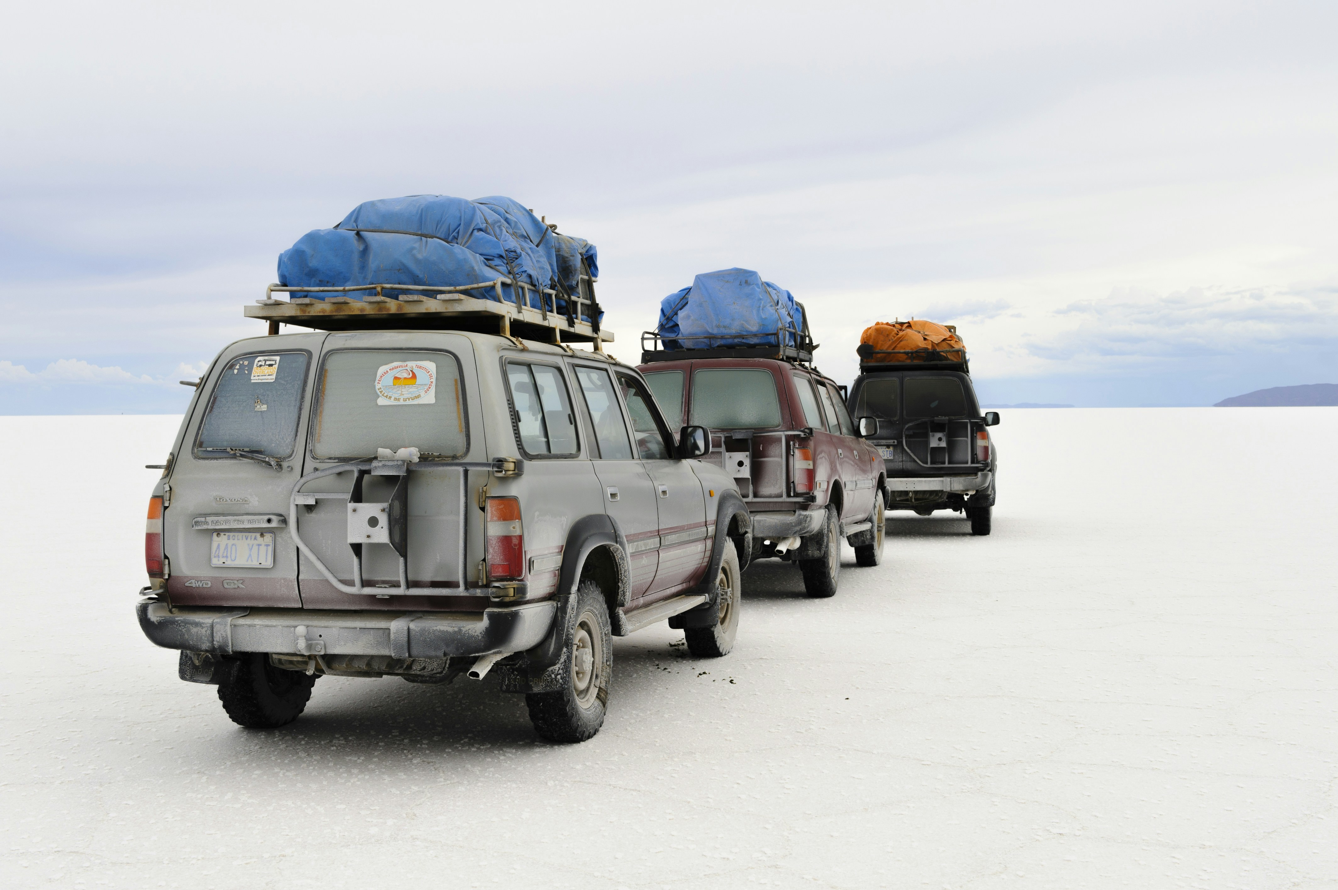 Three SUVs with rooftop luggage traverse the vast, white expanse of Bolivia's Uyuni Salt Flats under a cloudy sky.
