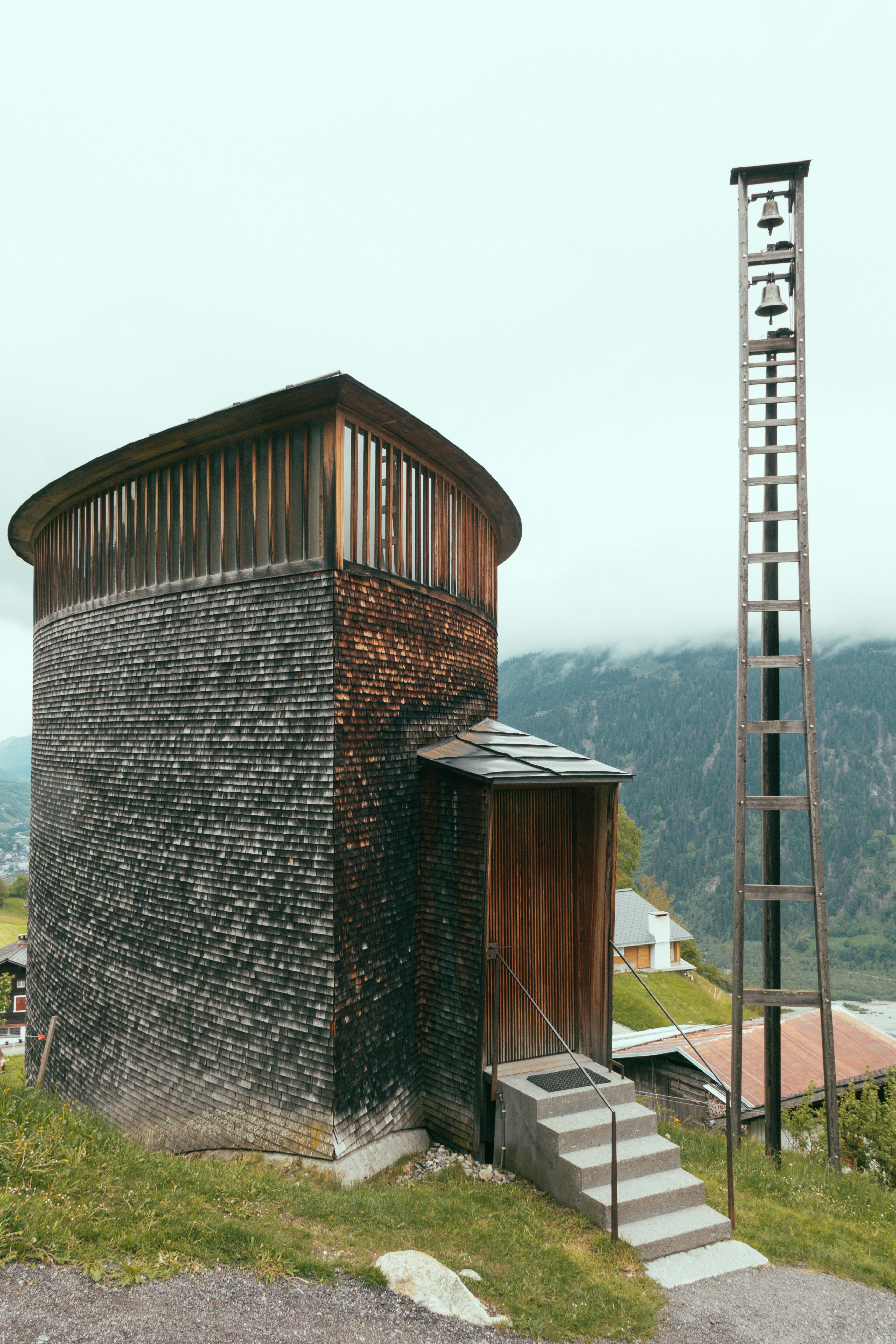 A uniquely designed wooden structure with a bell tower, nestled in a verdant landscape, showcasing a blend of modern architecture and rustic charm.