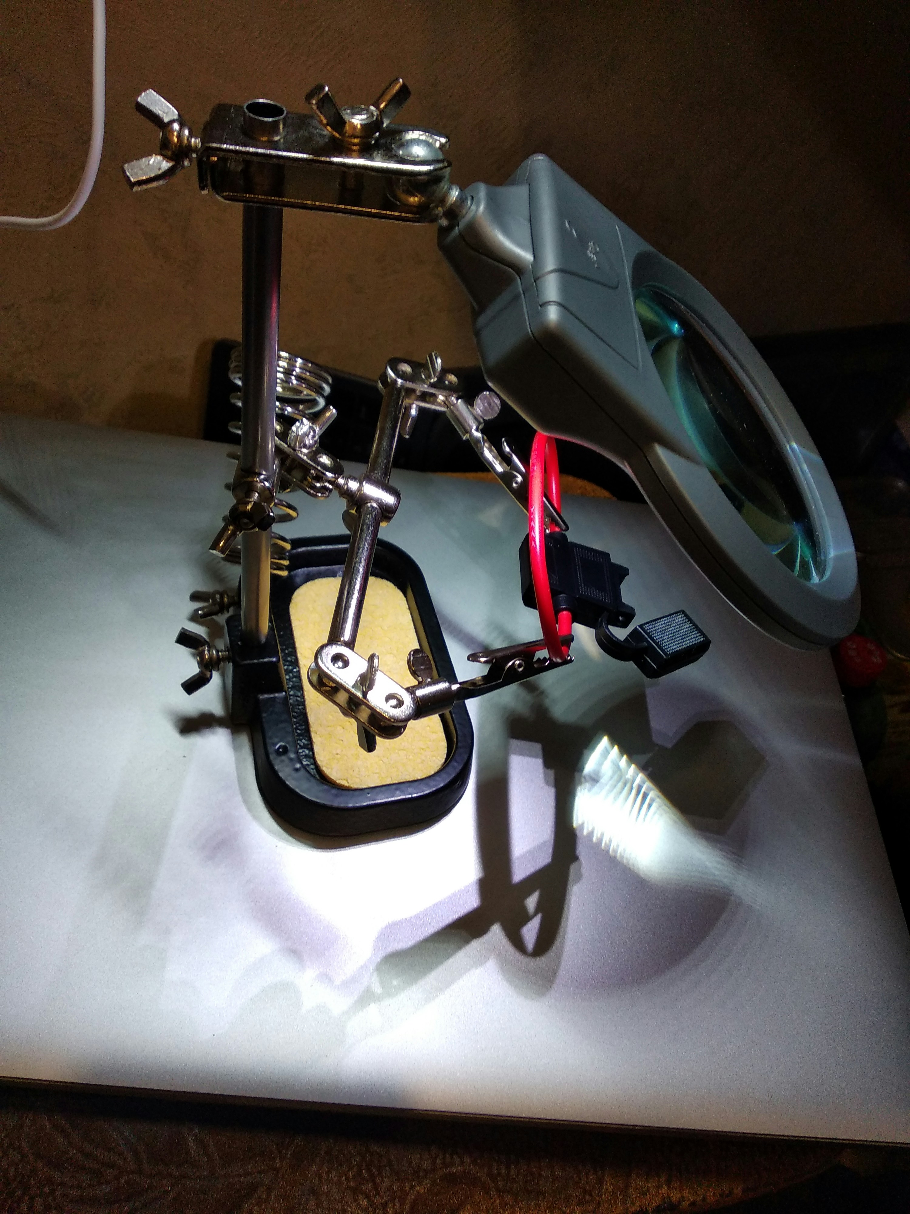 A magnifying glass and soldering aid positioned on a reflective surface, highlighting intricate tools used for detailed work. The setup casts unique shadows that enhance the composition.