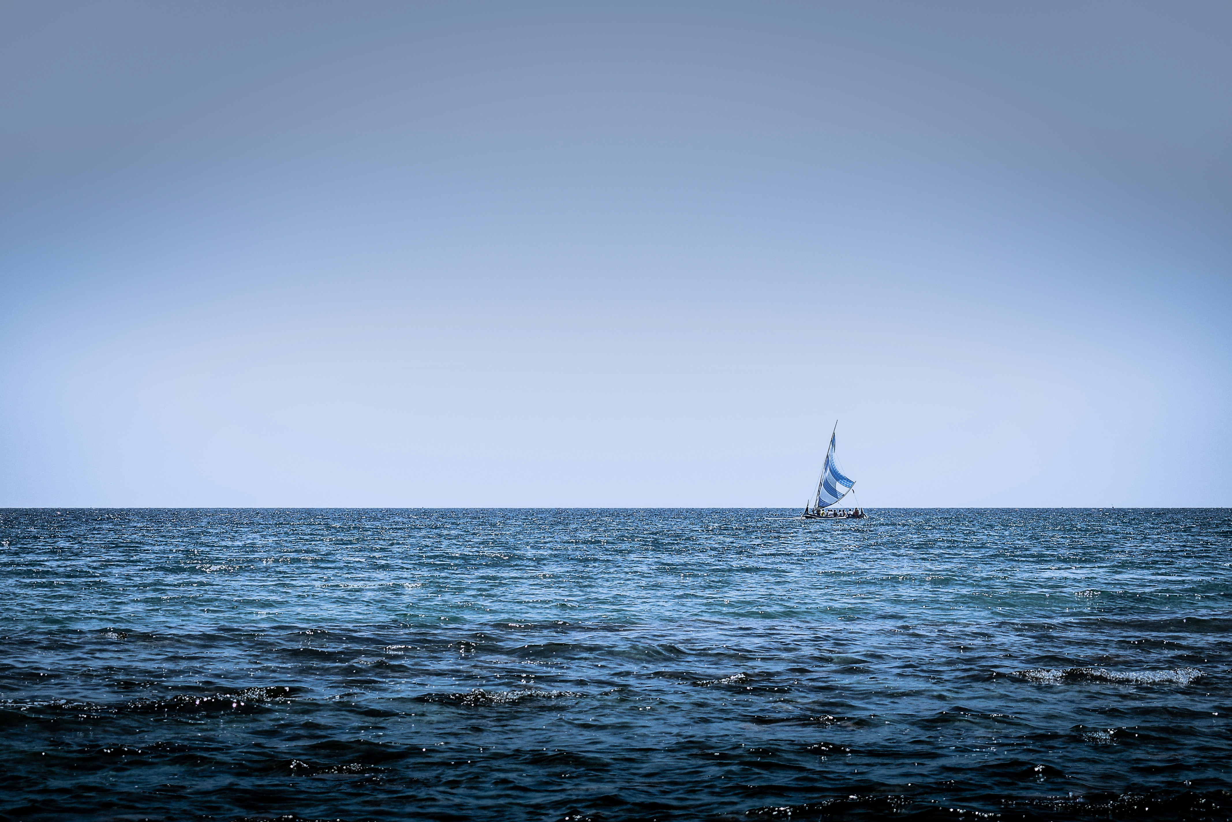 A lone sailboat glides across the calm sea, framed by a vast expanse of blue sky and water. The tranquil scene evokes a sense of peace and solitude.