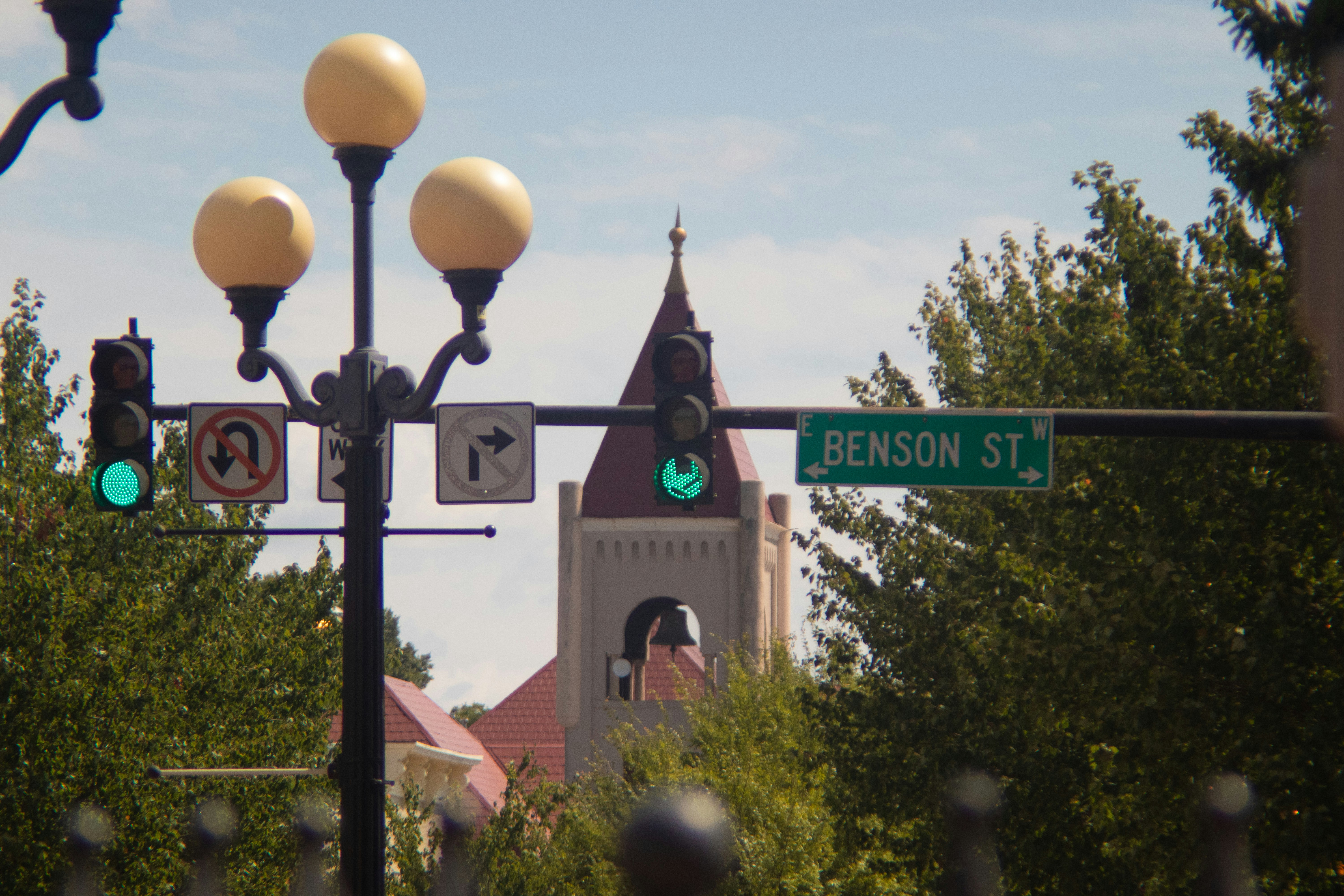 Traffic lights and a street sign for Benson St framed by greenery and architectural details in the background.