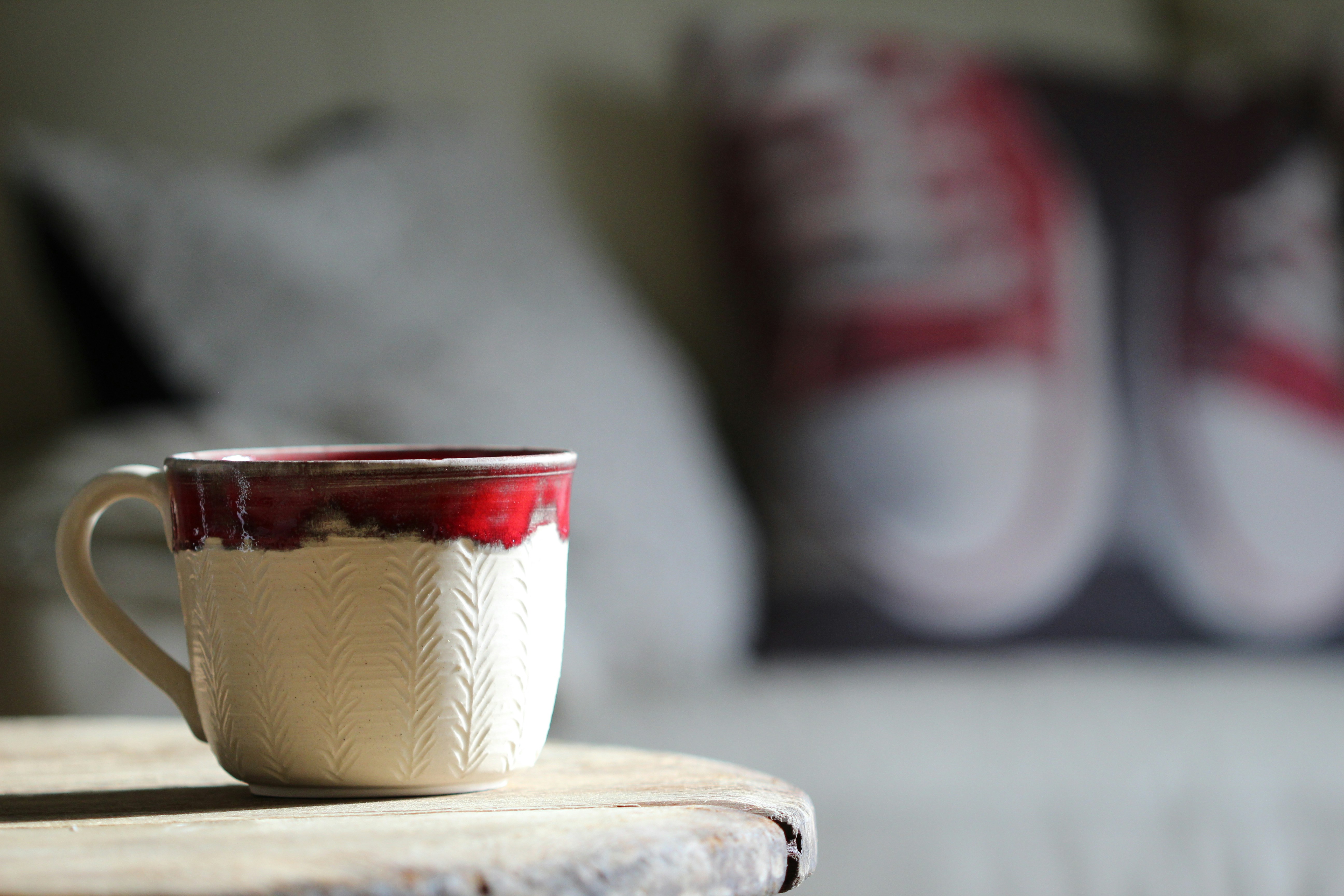 A textured ceramic mug rests on a rustic table, with a blurred background featuring a pair of sneakers on a cushion. The composition highlights the mug's intricate design.
