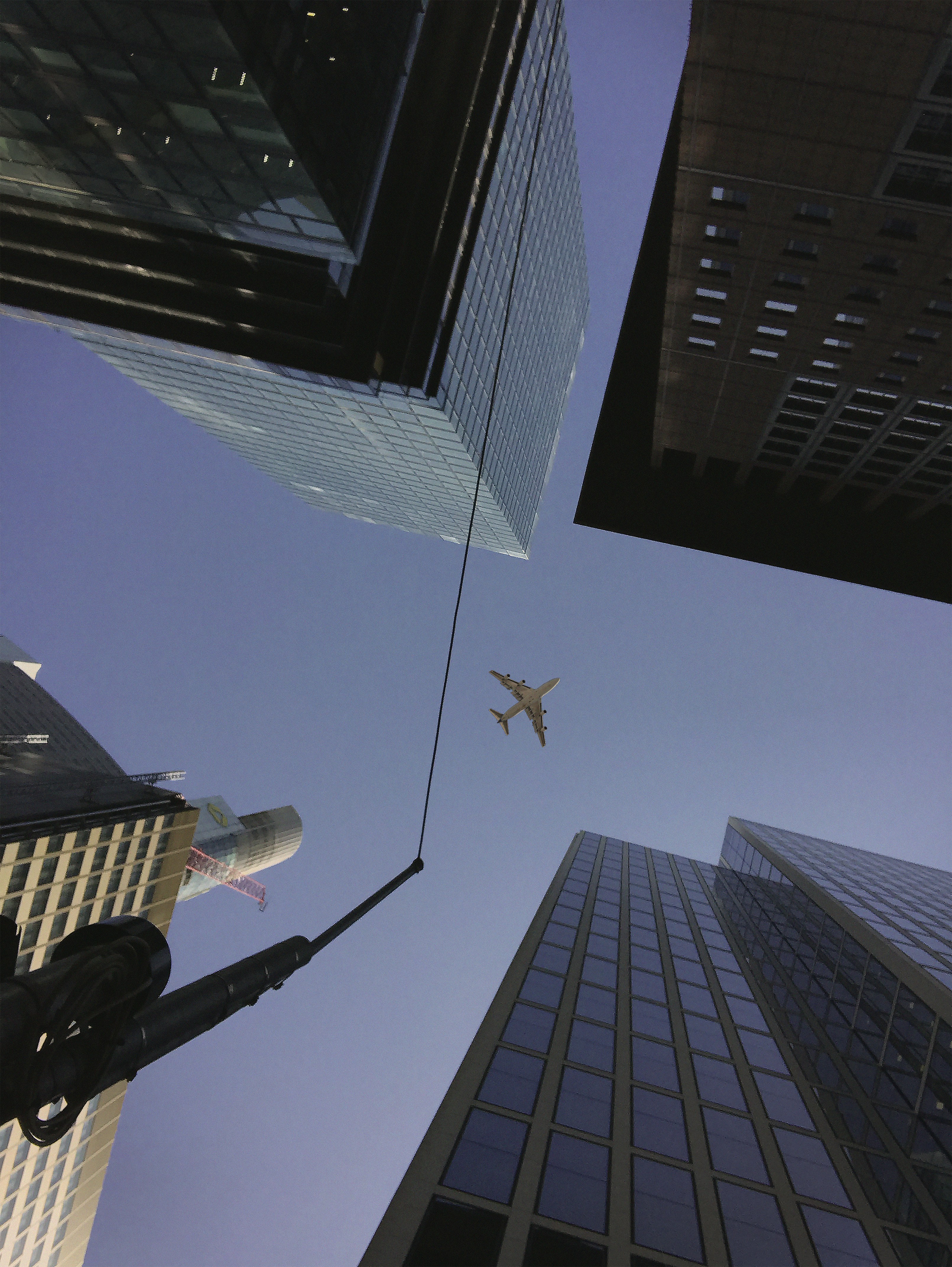 An airplane soaring above towering skyscrapers, captured from a unique low-angle perspective. The composition highlights the contrast between urban structures and the open sky.