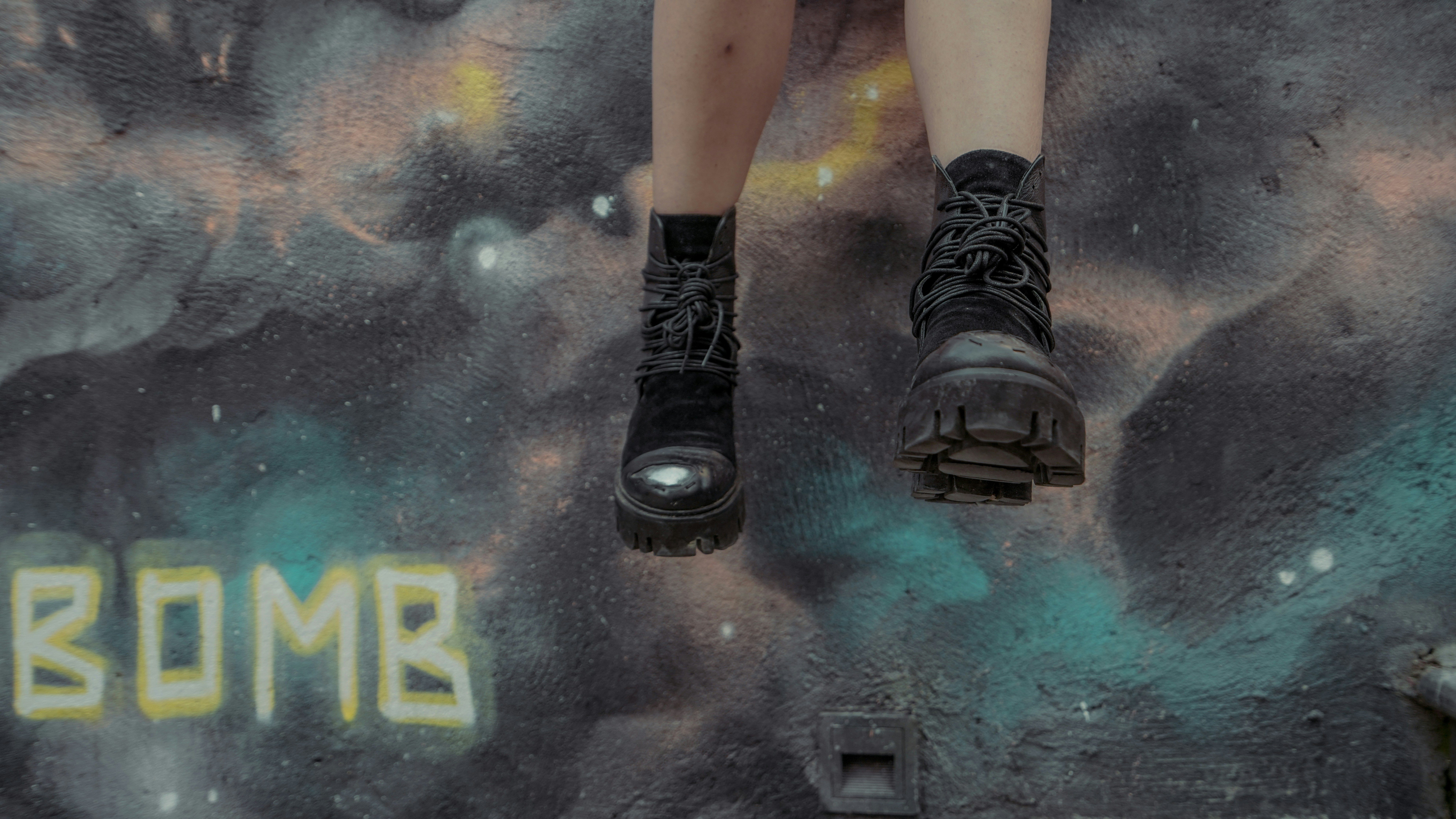 Black combat boots suspended above vibrant graffiti art featuring the word 'BOMB'.