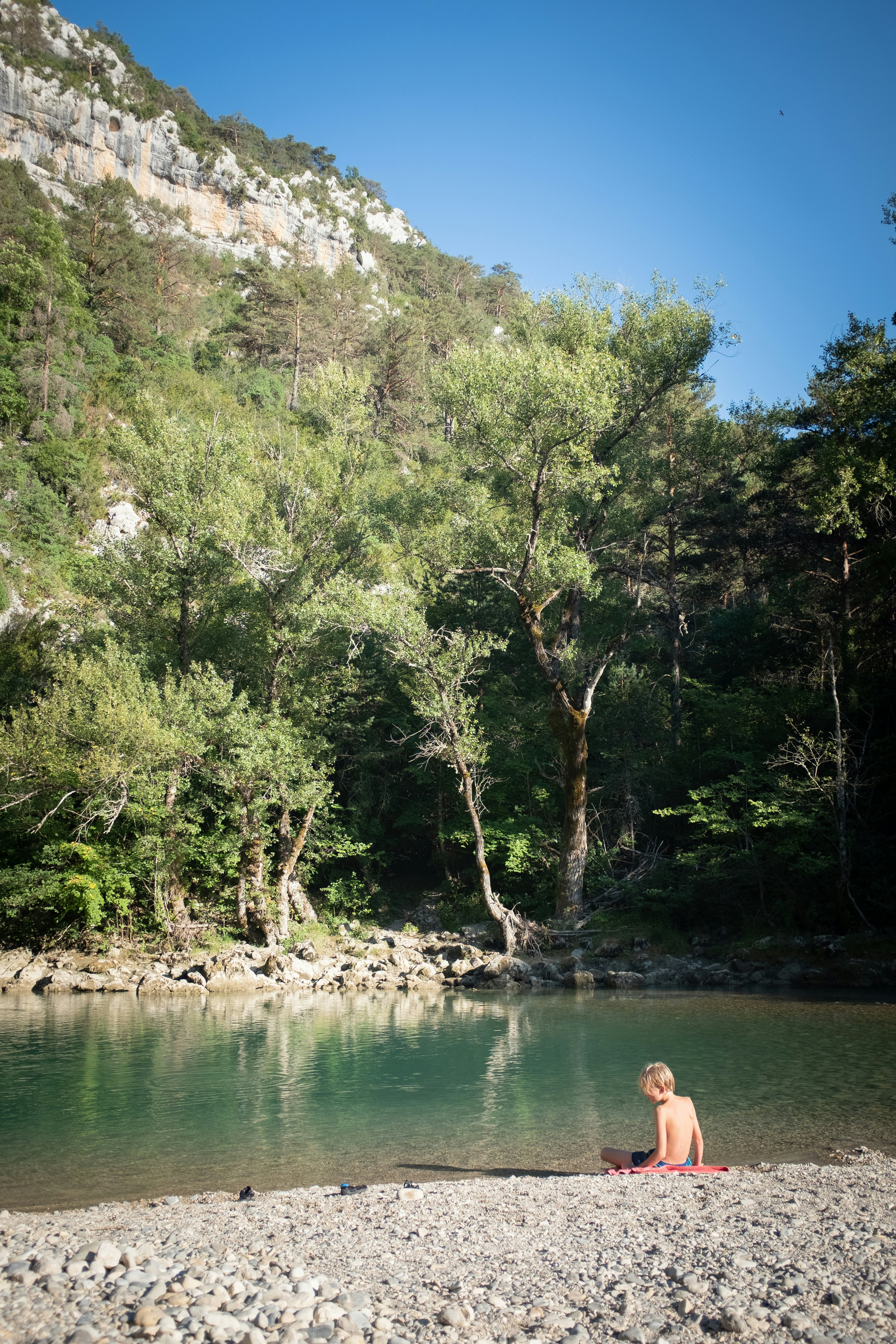 Sunlit Riverside Moment: a child at the water's edge beneath a limestone cliff