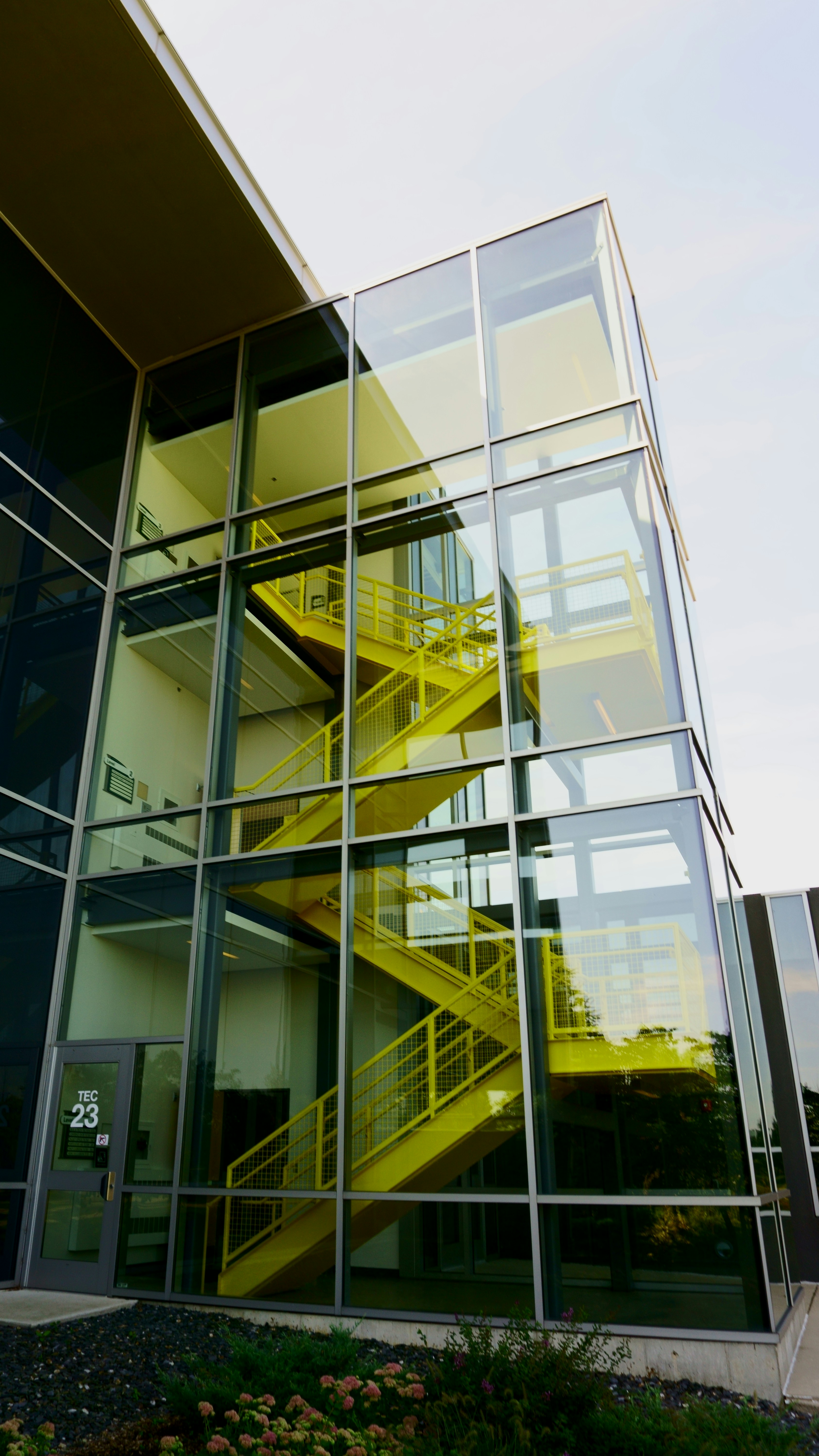 A modern building facade showcasing a yellow spiral staircase through reflective glass panels, emphasizing architectural design and color contrast.