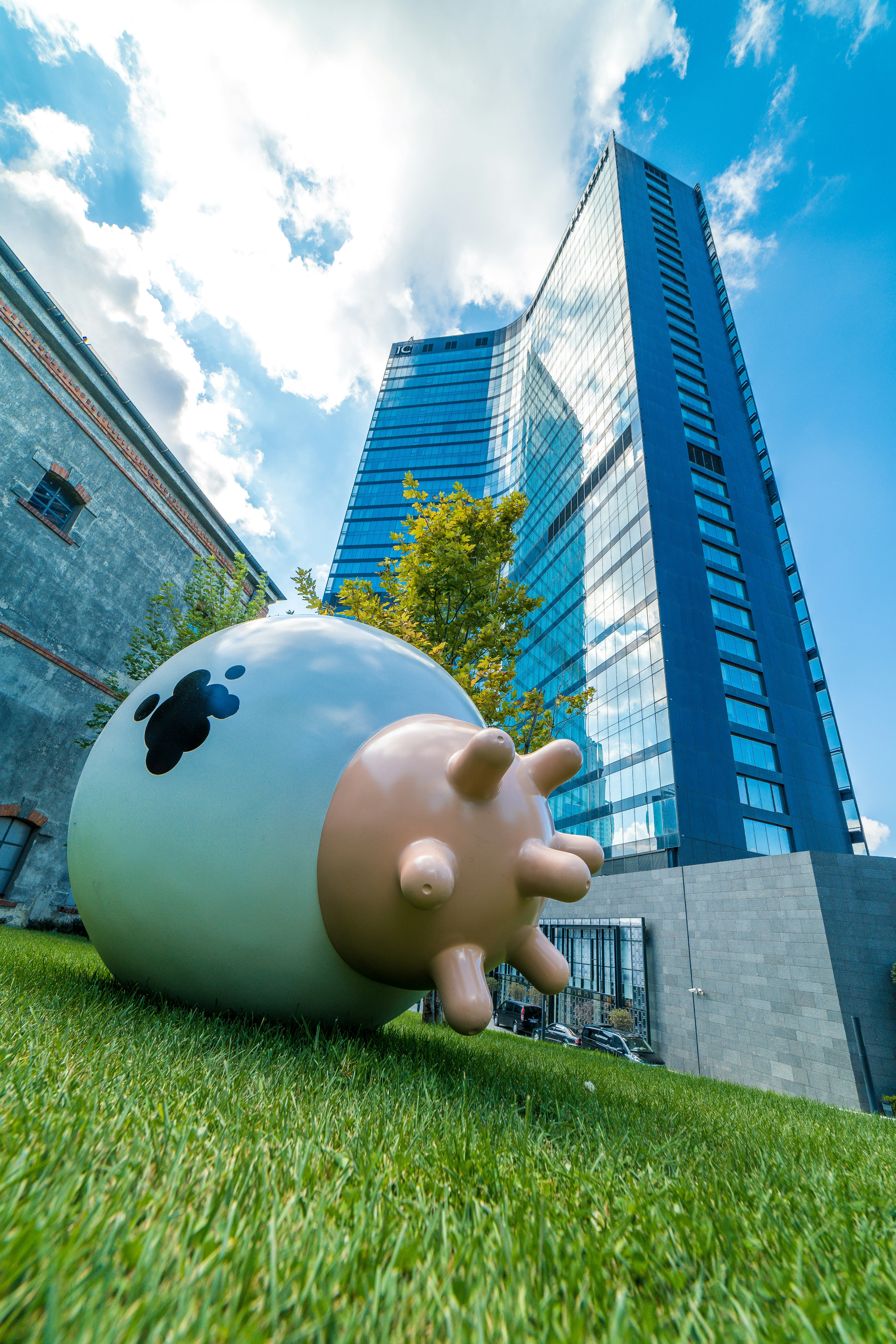 A playful sculpture resembling a cow's udder rests on lush grass, juxtaposed against a modern glass skyscraper and a historic building.