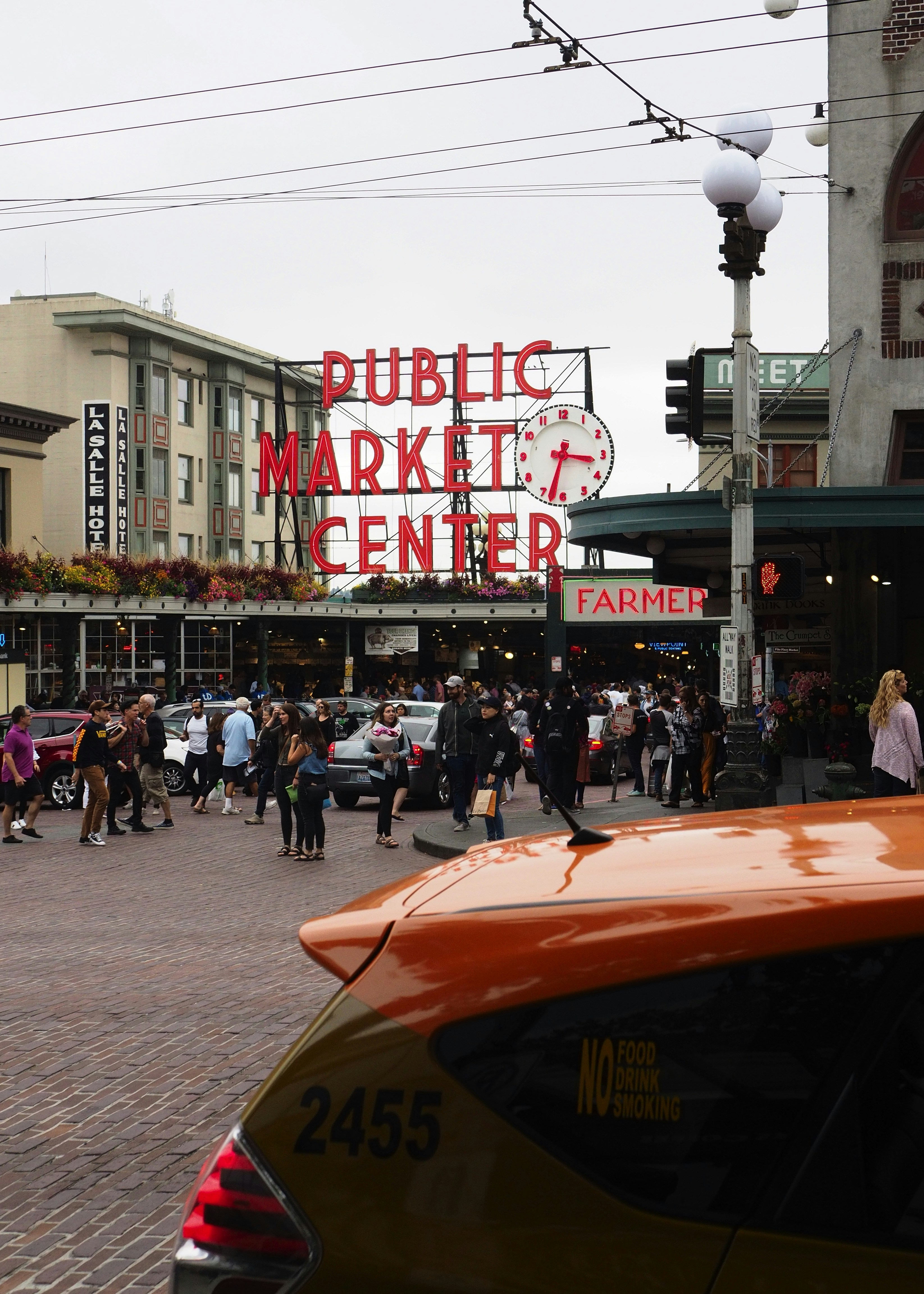 Public Market Giant: Neon Sign Over a Busy Plaza with a Bright Orange Vehicle in the Foreground
