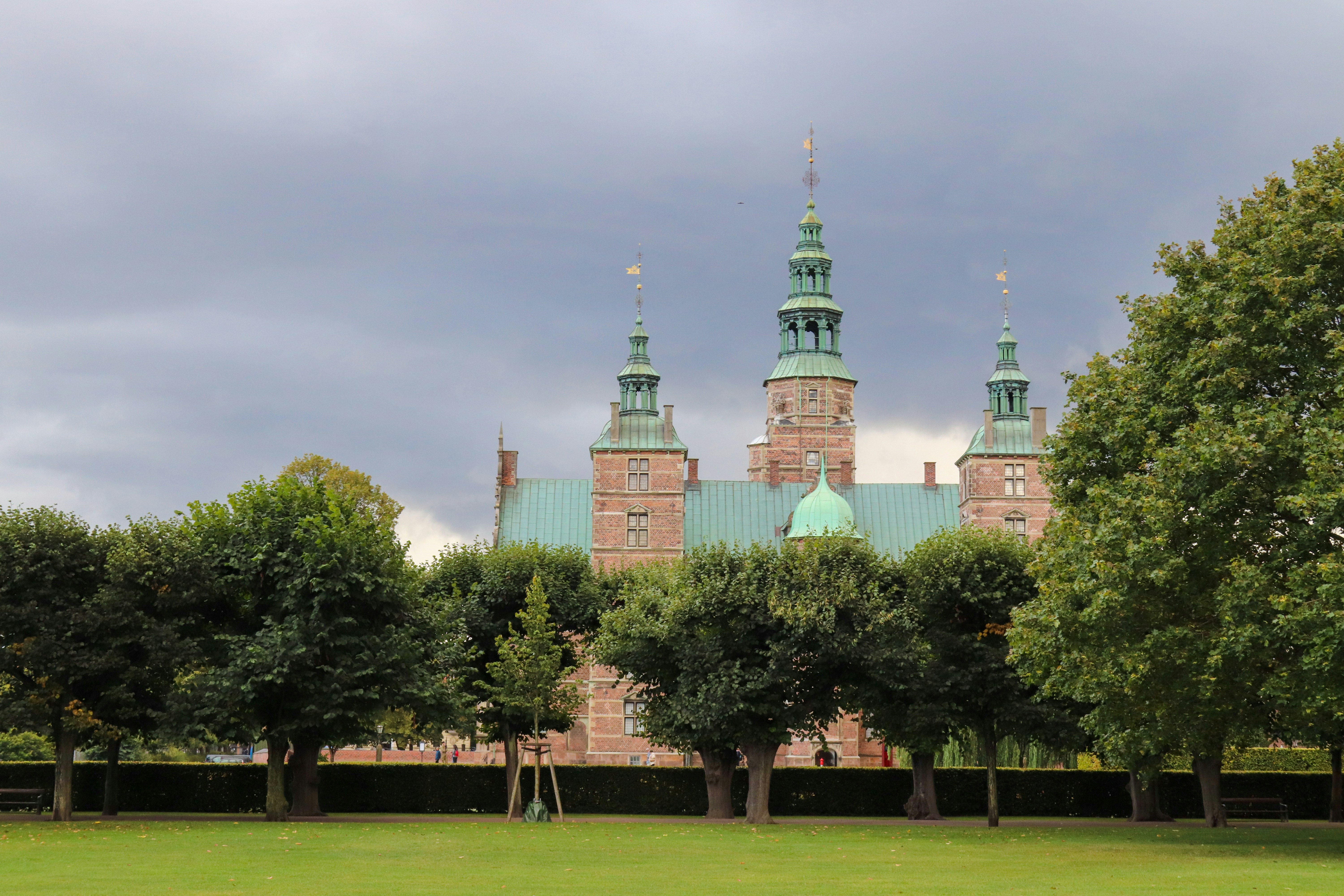 Rosenborg Castle stands majestically behind lush trees, showcasing its distinct architectural style against a moody sky.