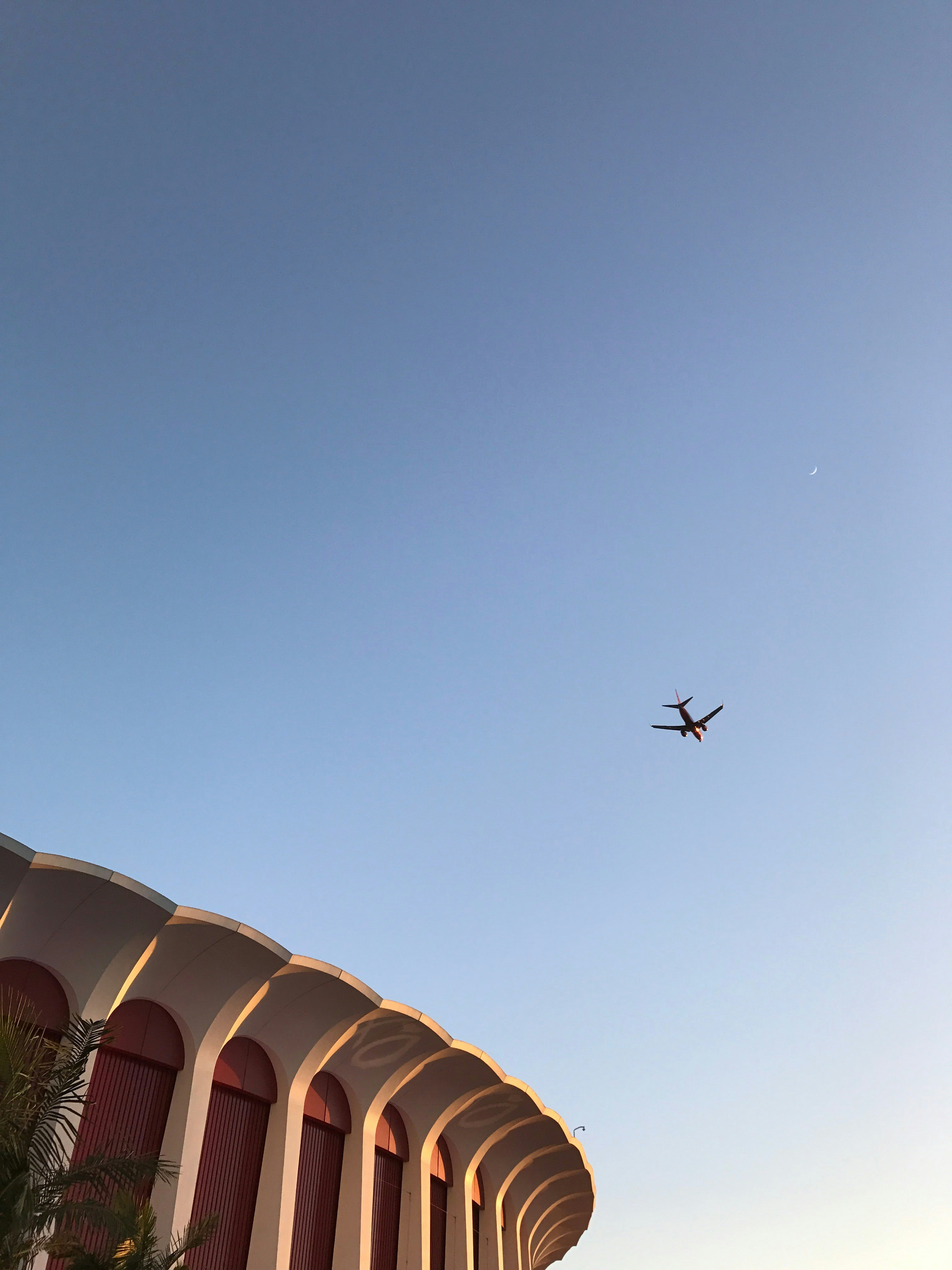 An airplane glides through a clear sky above a modern building with distinct curved architecture. The scene captures the harmony between man-made structures and aerial travel.