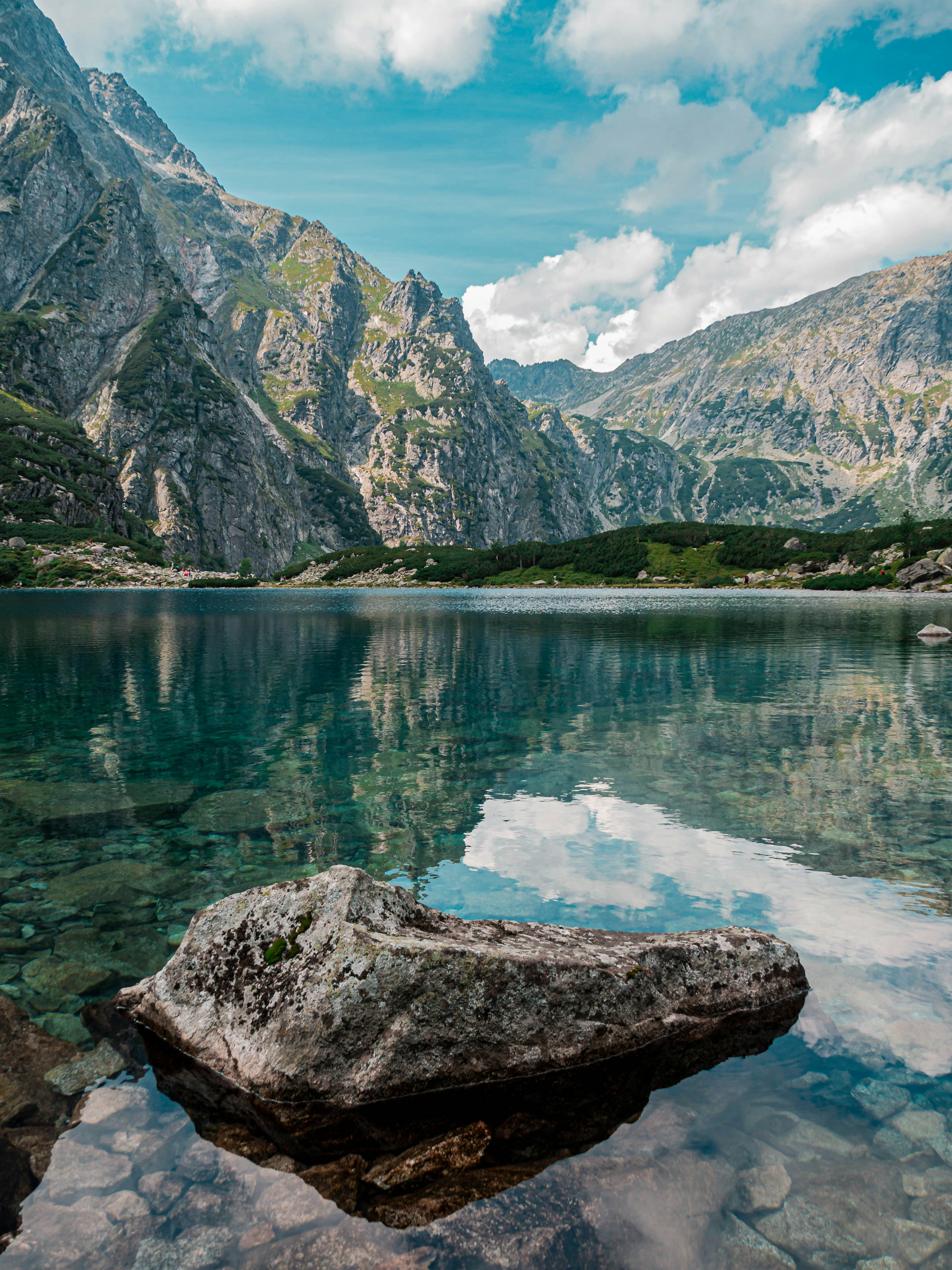 Clear mountain lake reflecting rugged peaks and fluffy clouds under a vibrant sky.