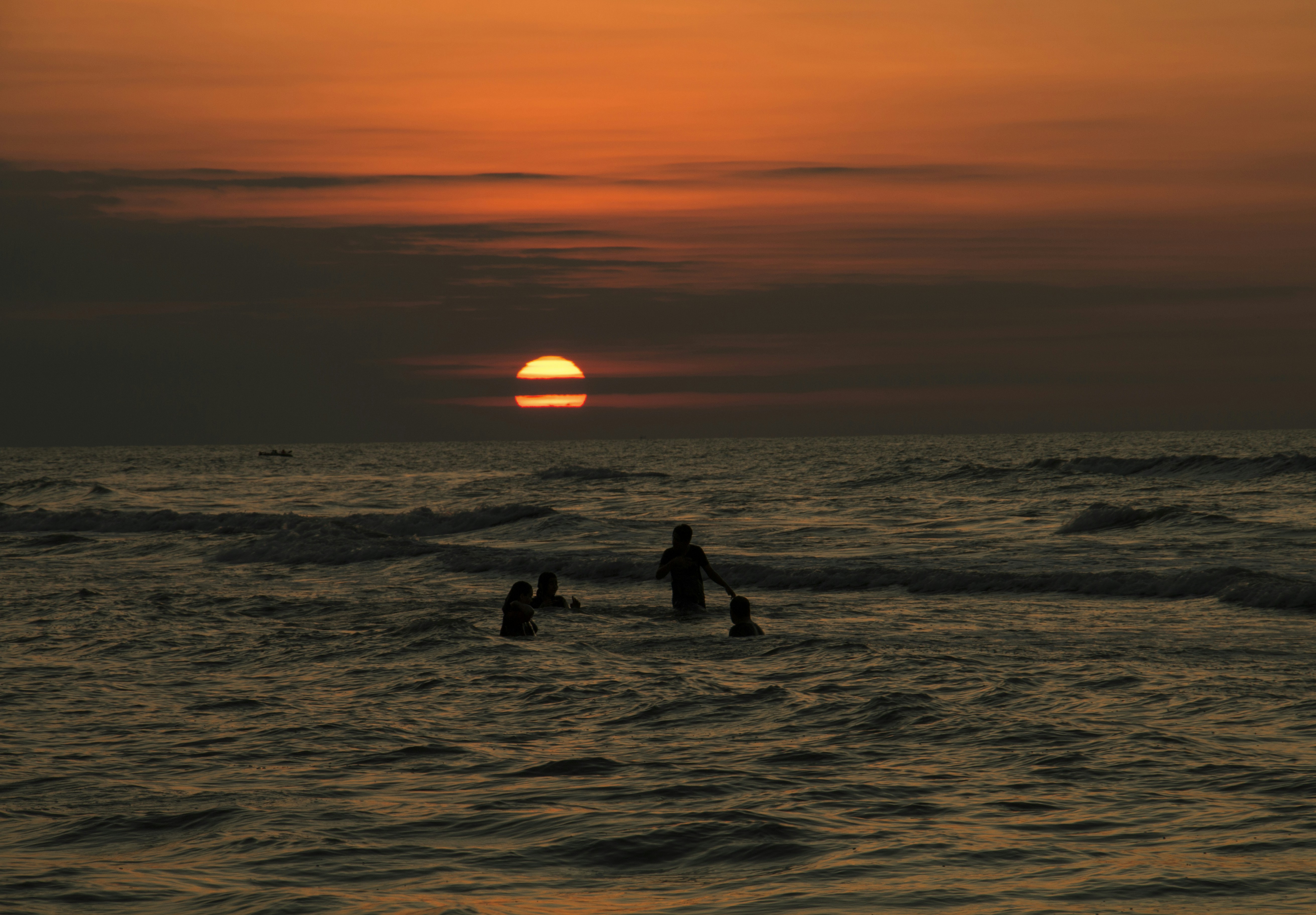 Silhouetted figures enjoying the ocean as the sun sets on the horizon, casting a warm glow over the water.