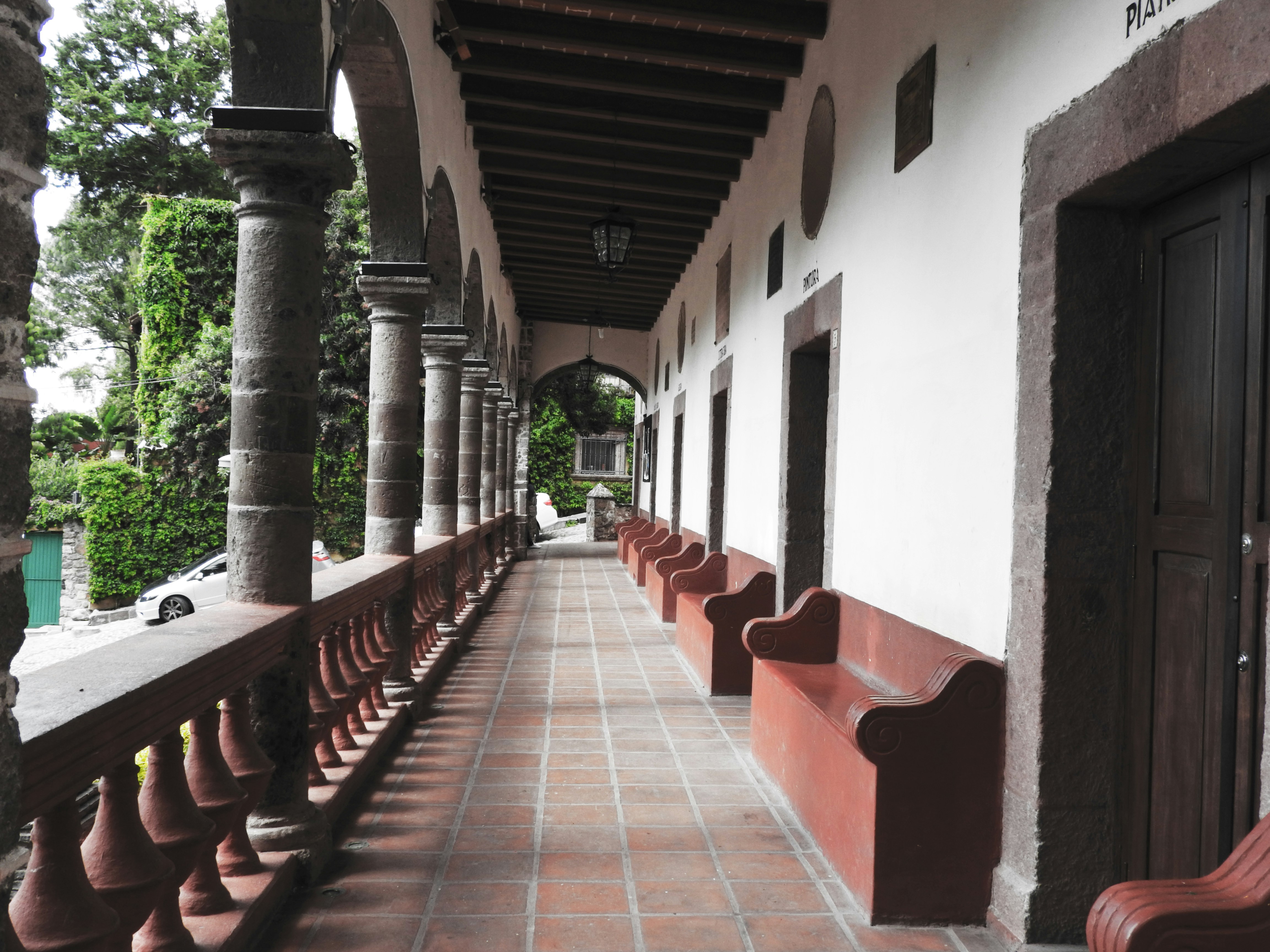 A tranquil corridor featuring rustic wooden benches and stone columns, framed by lush greenery outside. The architectural details evoke a sense of timelessness.