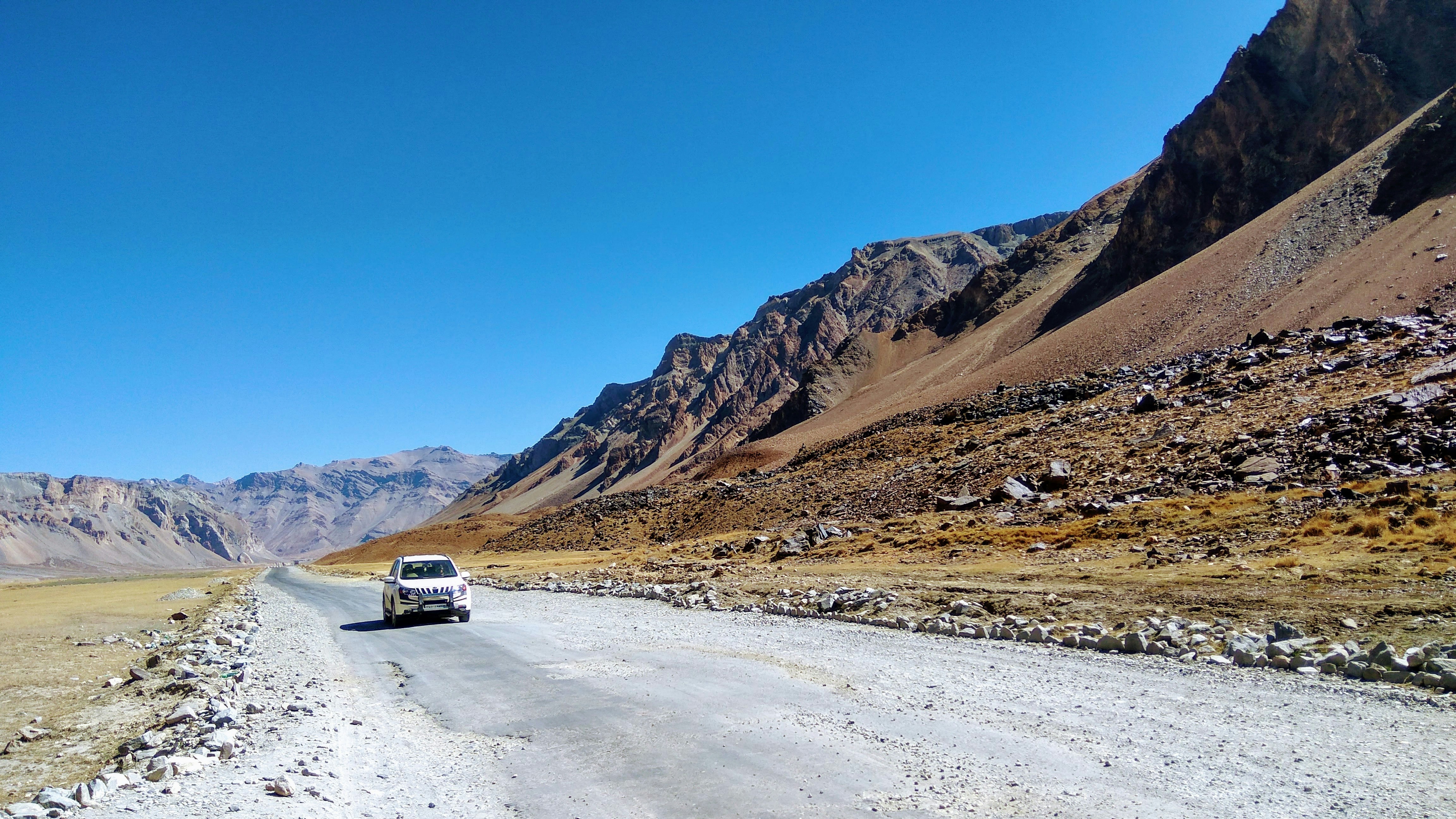 A white SUV travels a gravel road through a barren mountain landscape under a bright blue sky.