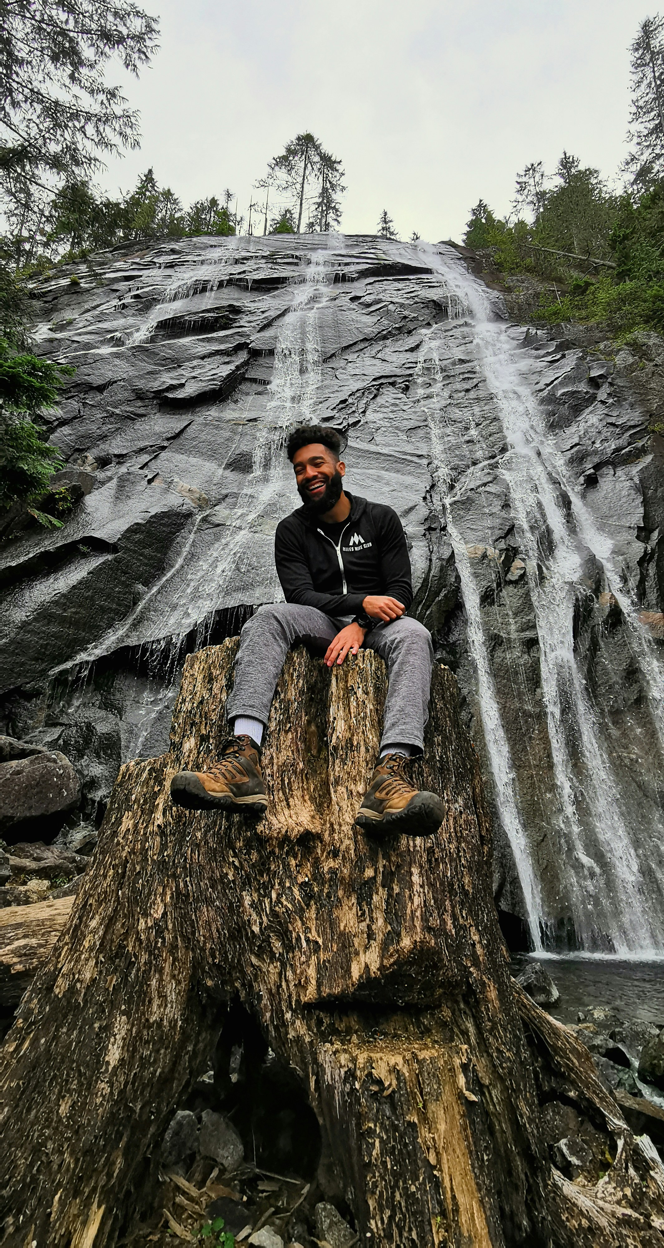 Person seated on a tree stump in front of a cascading waterfall surrounded by lush greenery.